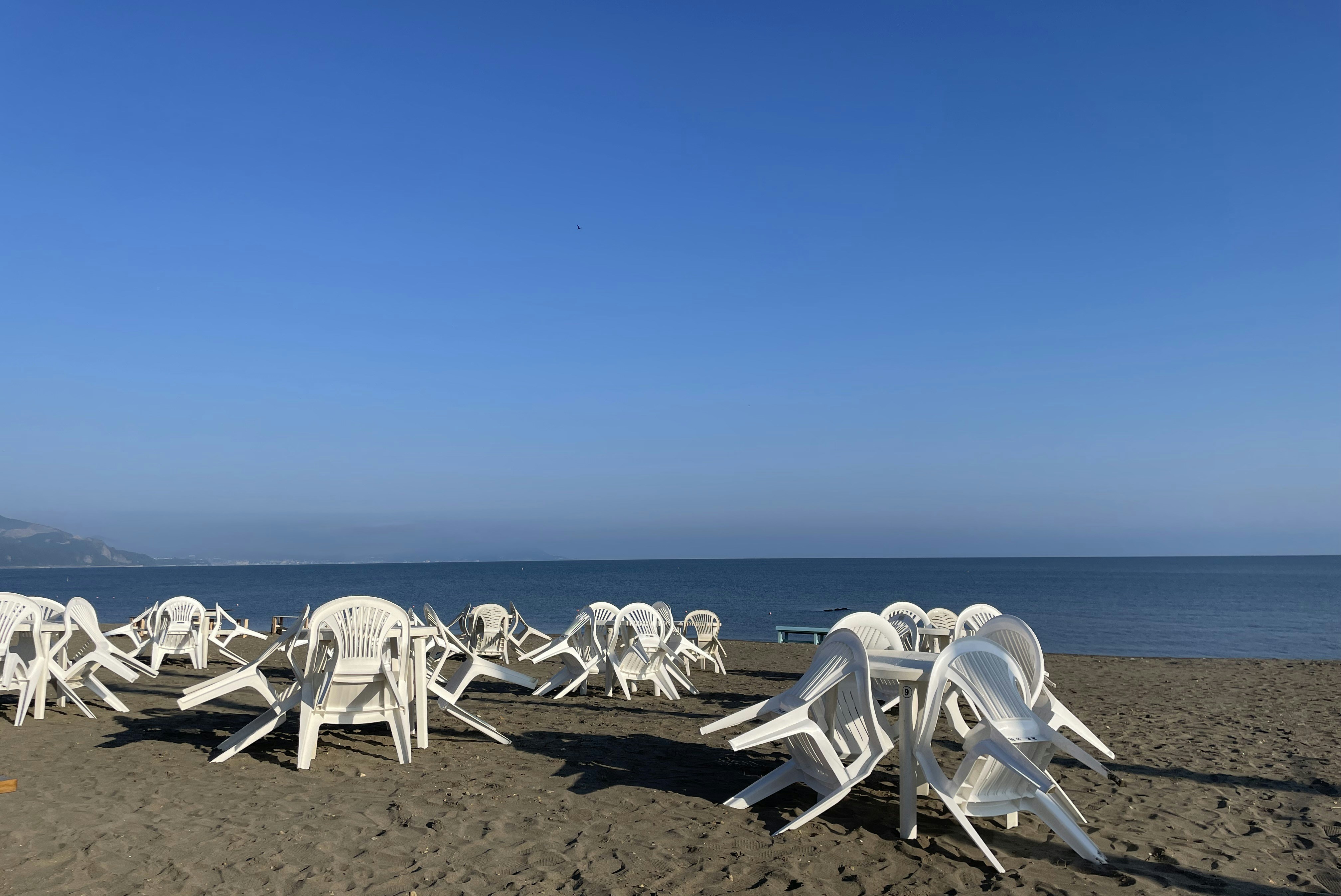 White plastic chairs scattered along a sandy beach with calm sea and clear blue sky in the background. A simple, serene setup without people, suggesting a paused moment or event readiness.
