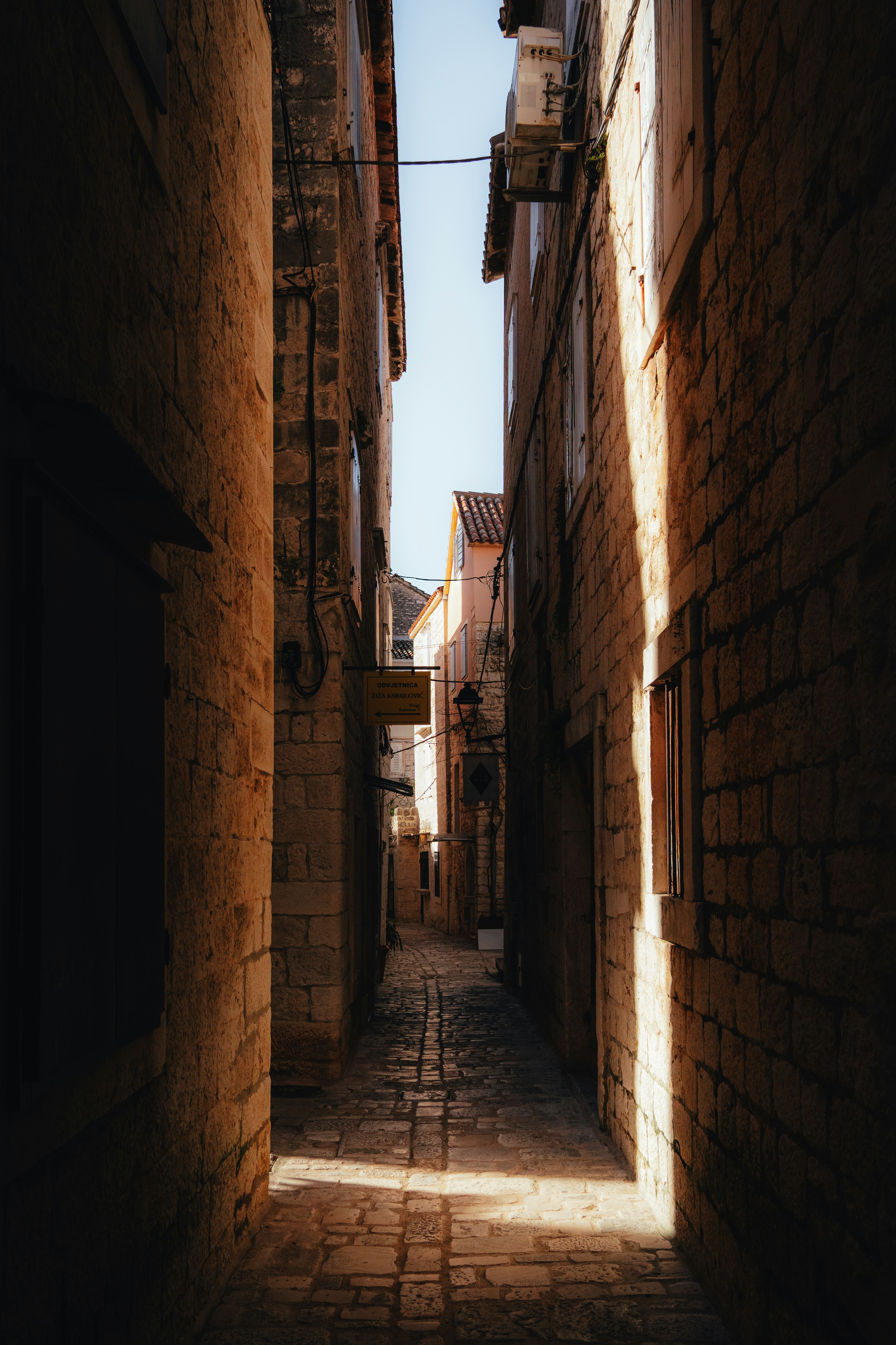 A sunlit alley framed by weathered stone walls, with a narrow cobblestone path leading toward a bright opening at the far end. This is a photograph capturing a quiet, historic street scene.
