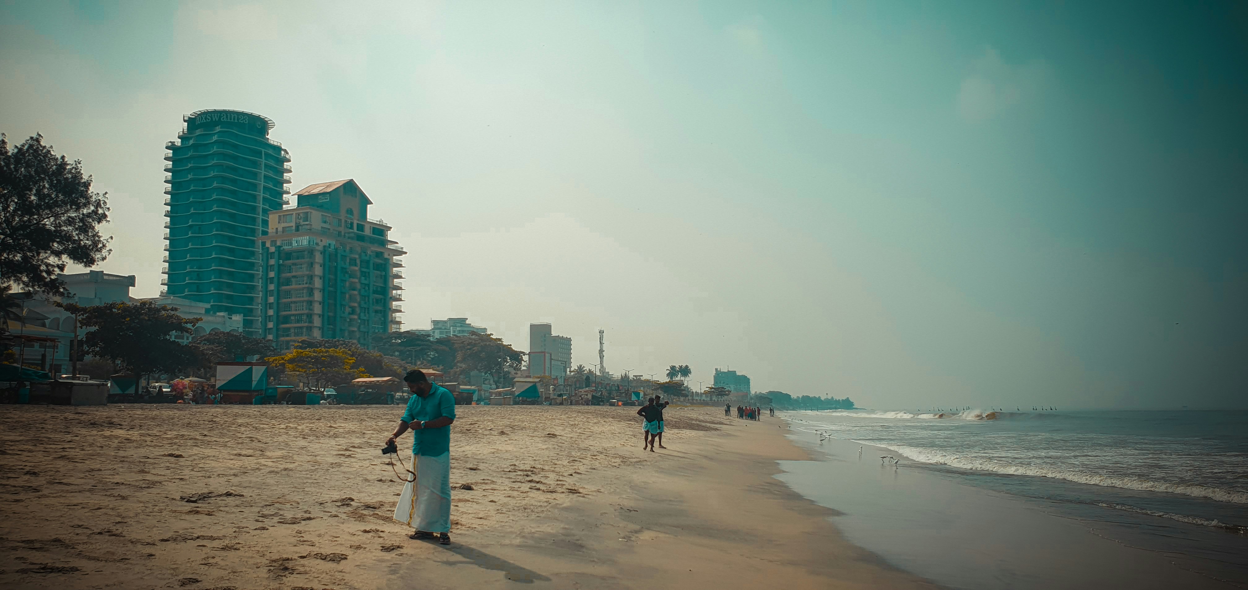 A wide landscape photograph capturing a sandy beach coastline with city high-rises in the background under a pale sky.