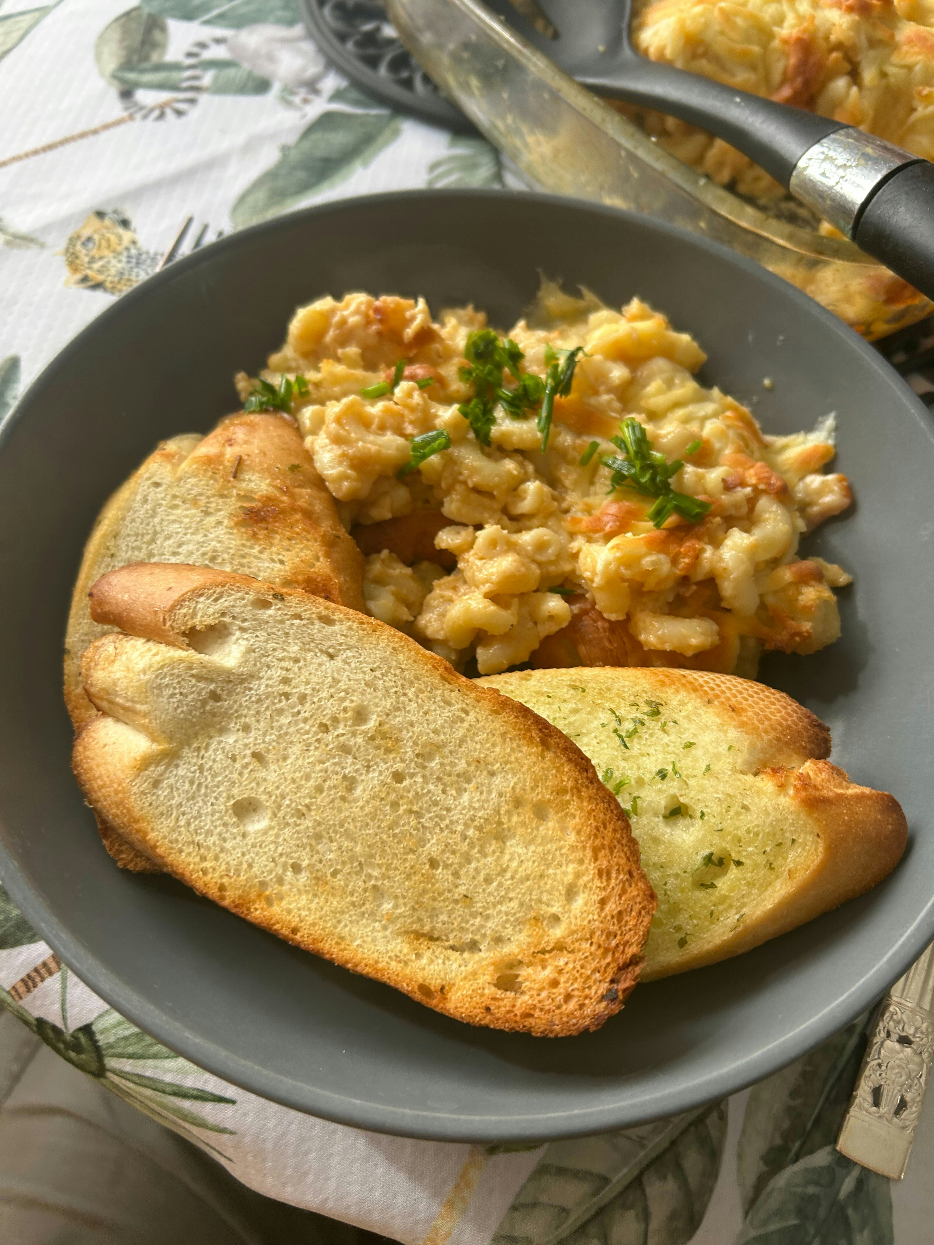 Creamy scrambled eggs with herbs on a grey plate, accompanied by toasted bread slices; a pan and utensils rest nearby, suggesting a home-cooked breakfast.