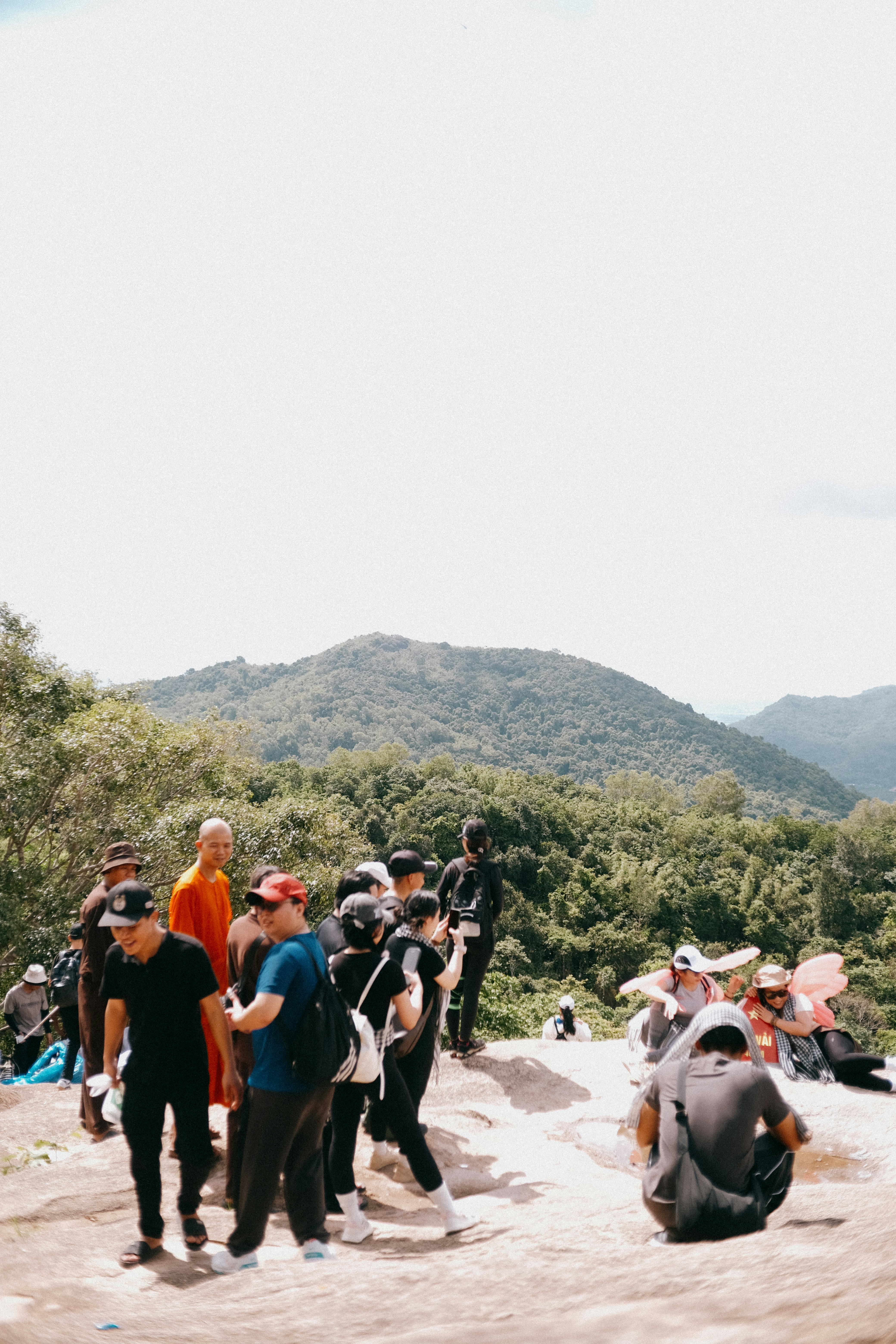 A group of hikers congregates on a sunlit rock ledge with a forested hillside and distant mountains in the background.
