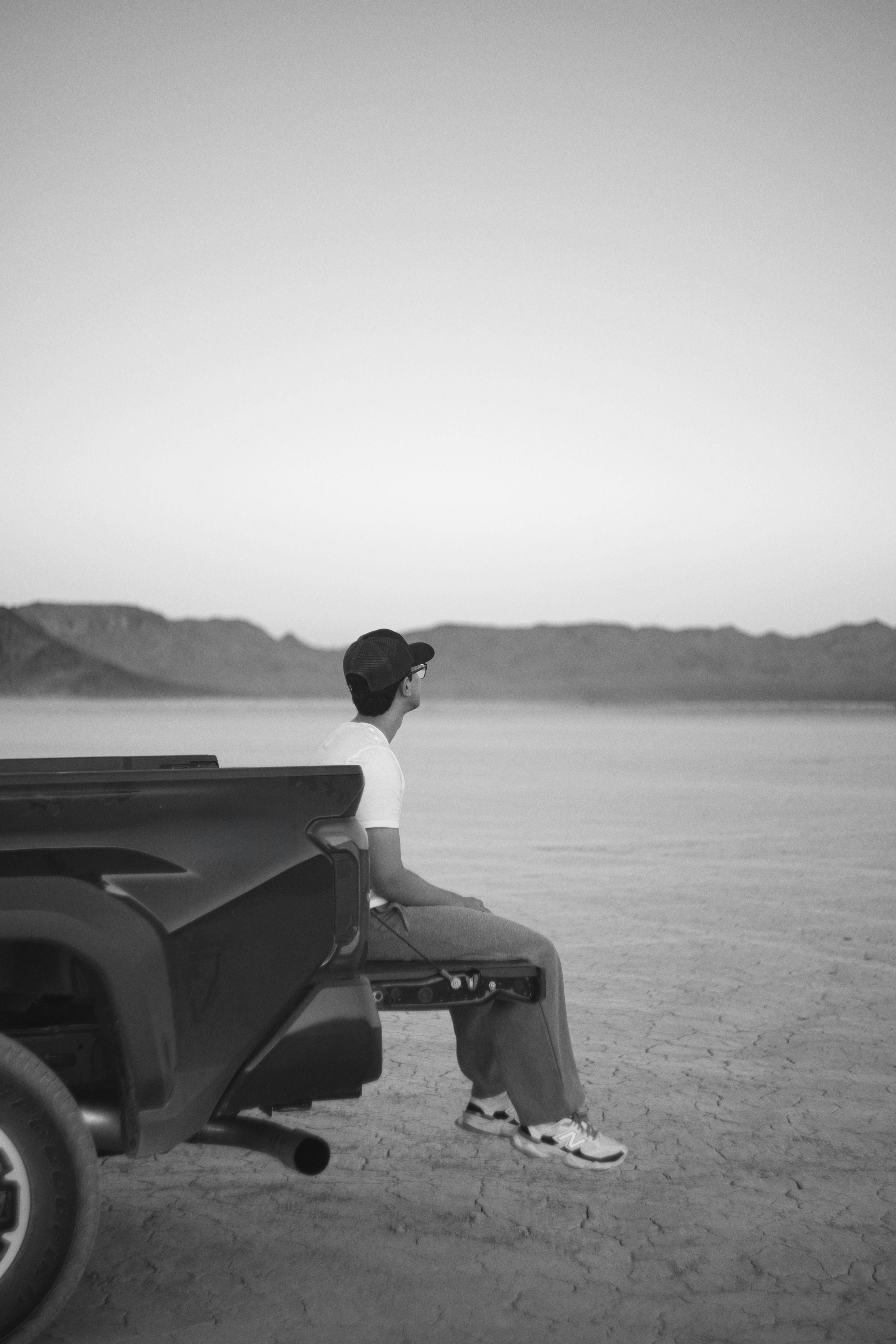 A man sits on the tailgate of a pickup facing a vast salt flat, mountains in the distance; grayscale emphasizes solitude and vastness of the landscape.