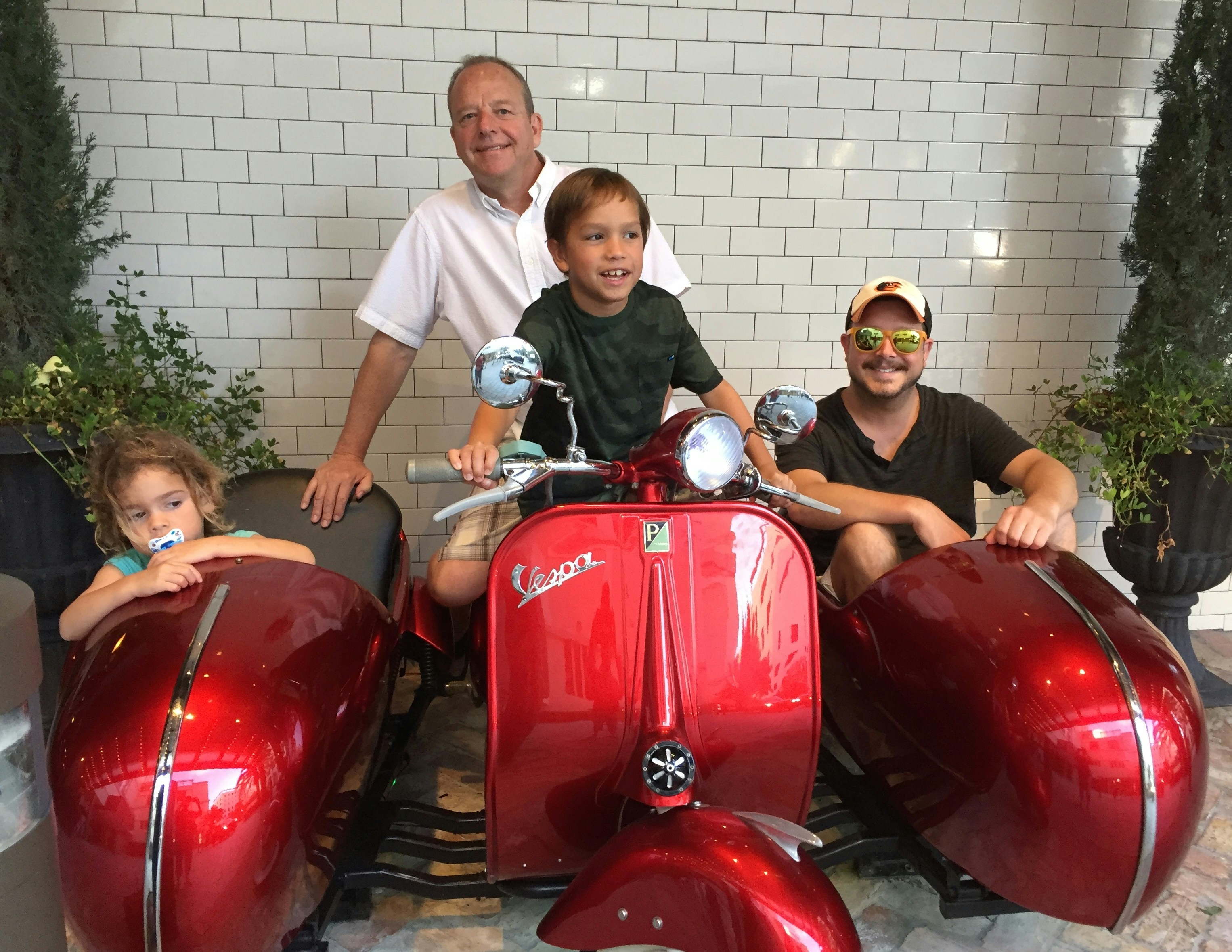 Three smiling children and two adults posing with a glossy red three-wheeled motorcycle at a showroom; chrome accents and tiled backdrop provide a bright, playful setting.