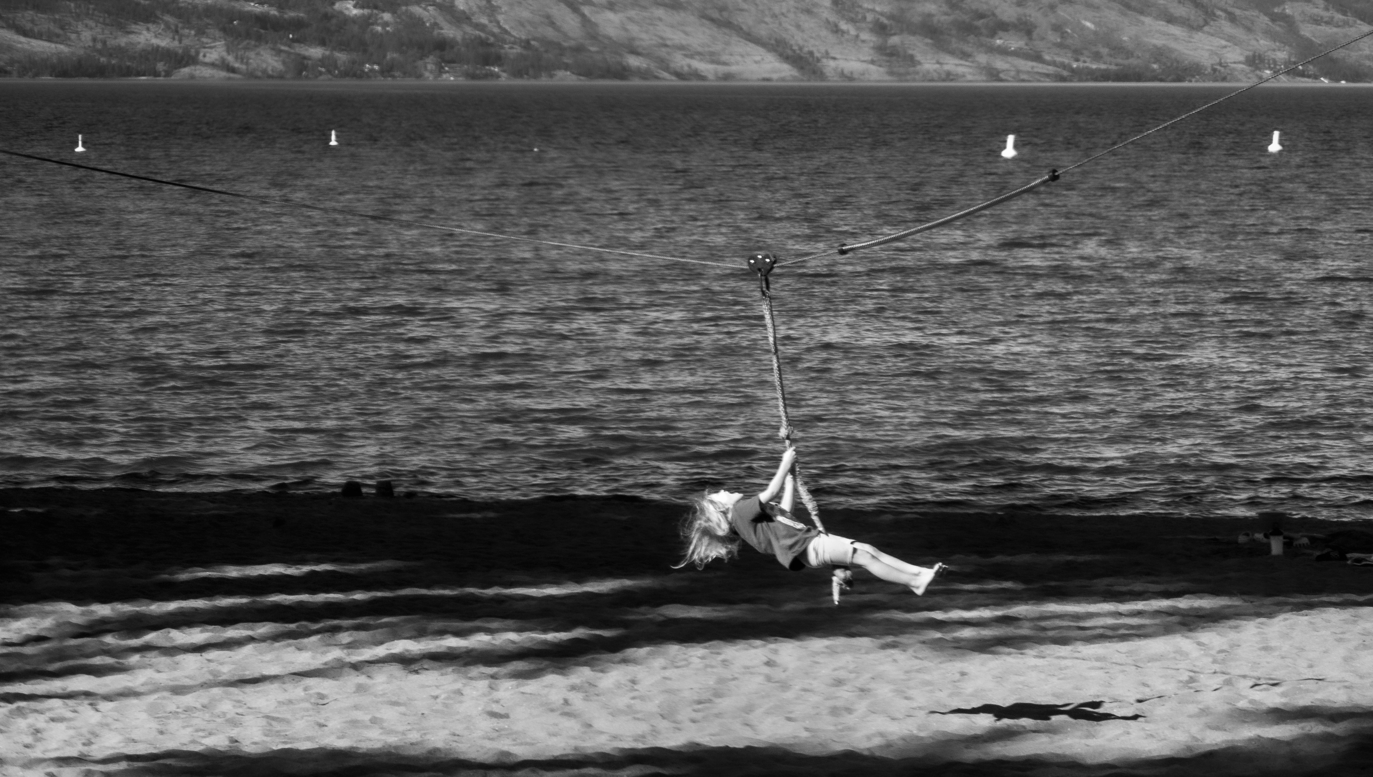 Child on a swing over the beach in subdued grayscale, with ocean and distant land in the background. Monochrome still emphasizing texture of sand, water, and figure.