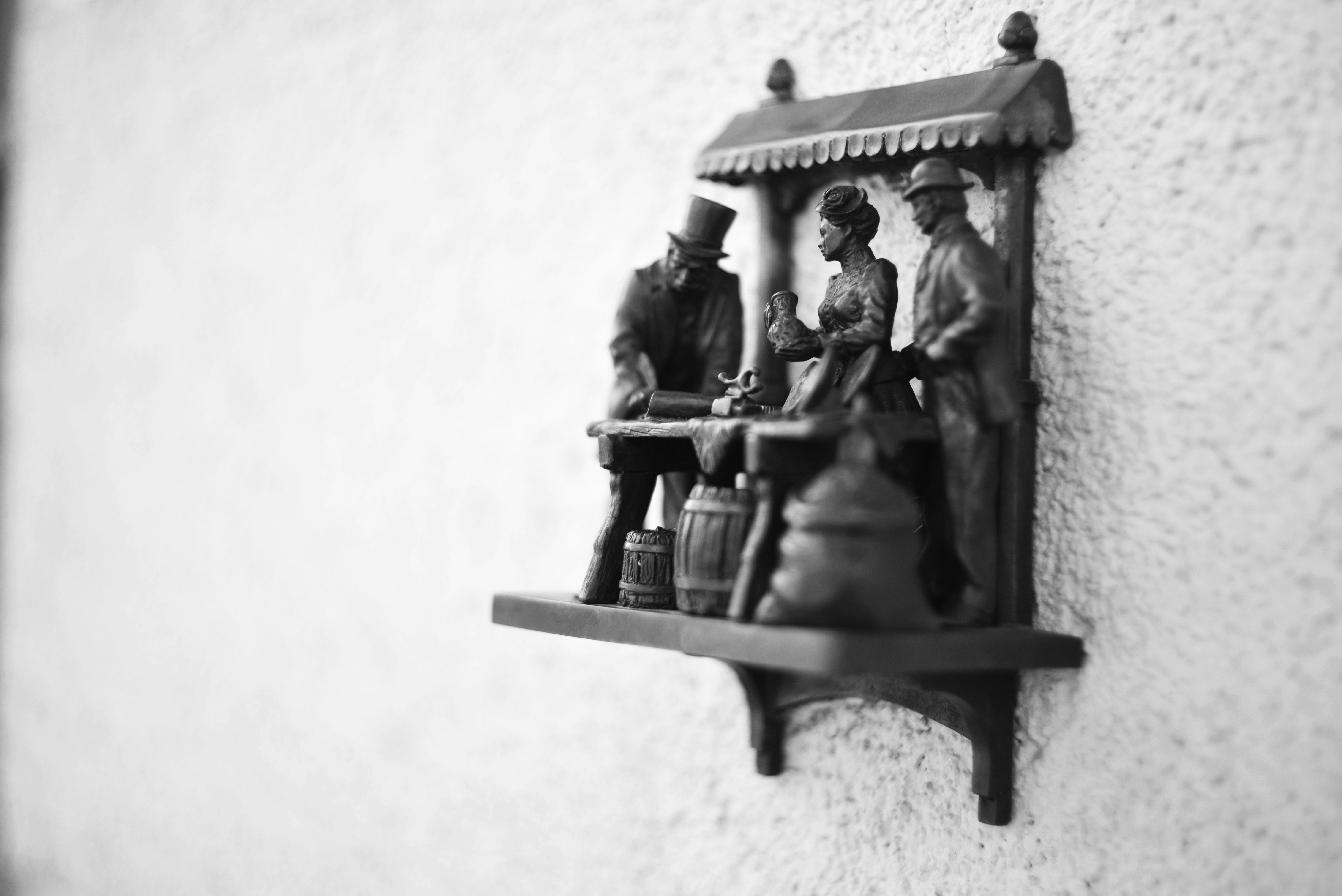 A small three-dimensional figurine scene mounted on a wall, captured in grayscale with shallow depth and a gently blurred backdrop.
