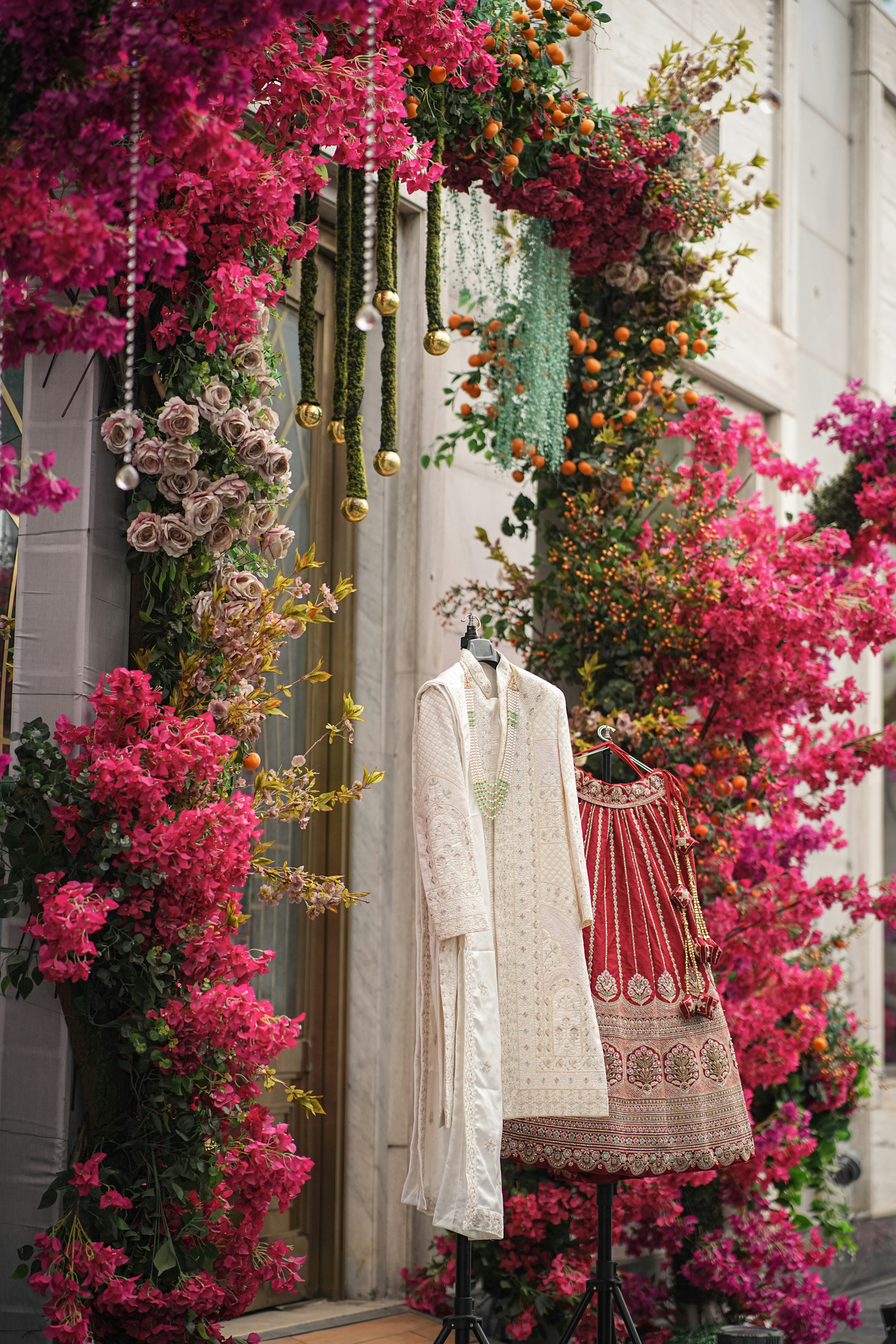 Colorful floral storefront with traditional garments on dress forms framed by vivid pink blossoms; a decorative display of clothing against a floral arch. Logo-like display of textiles stands out.