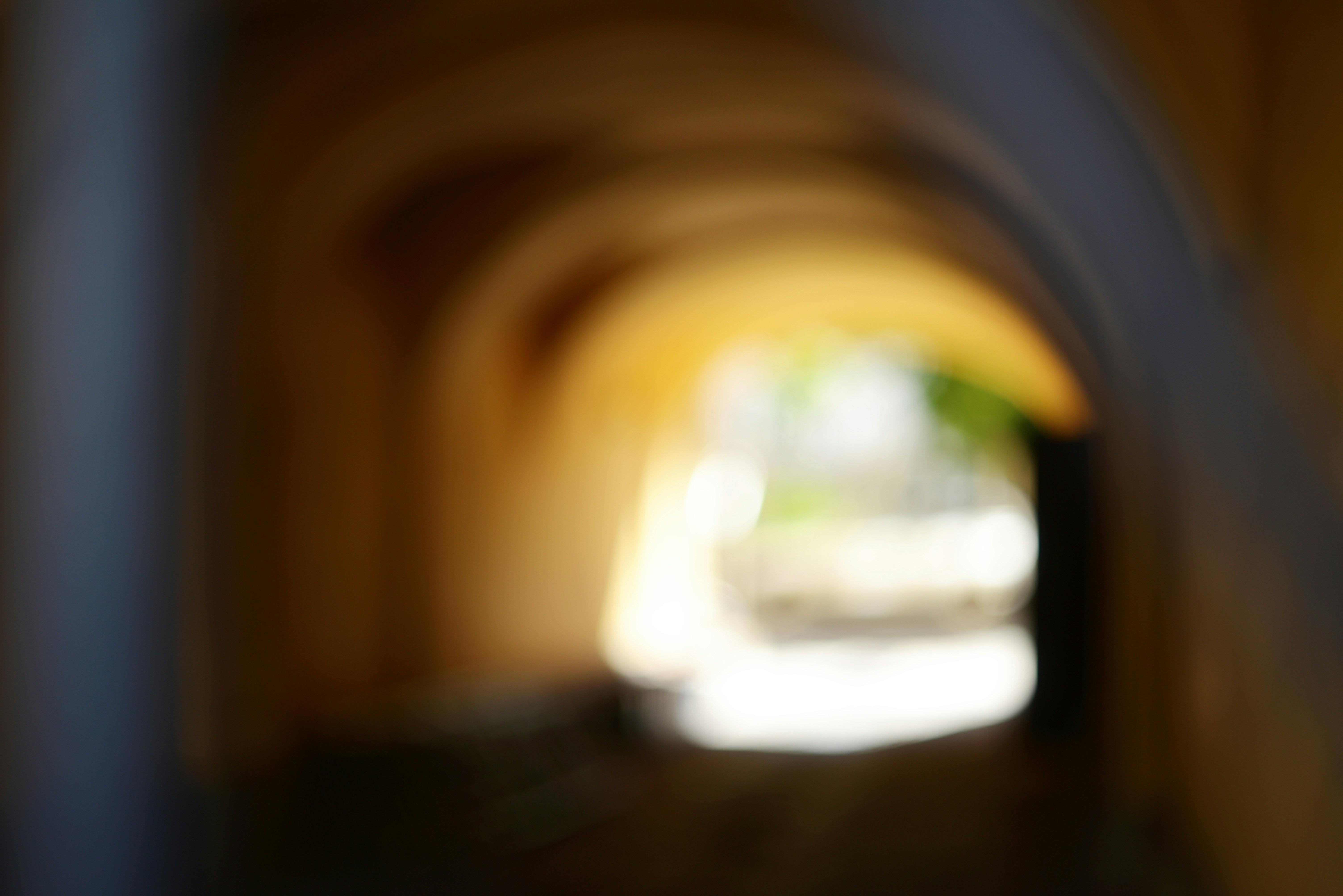 An abstract, out-of-focus interior scene with a bright opening at the end of a tunnel-like passage, creating a warm glow and soft bokeh. The image emphasizes mood and atmosphere rather than defined subjects.