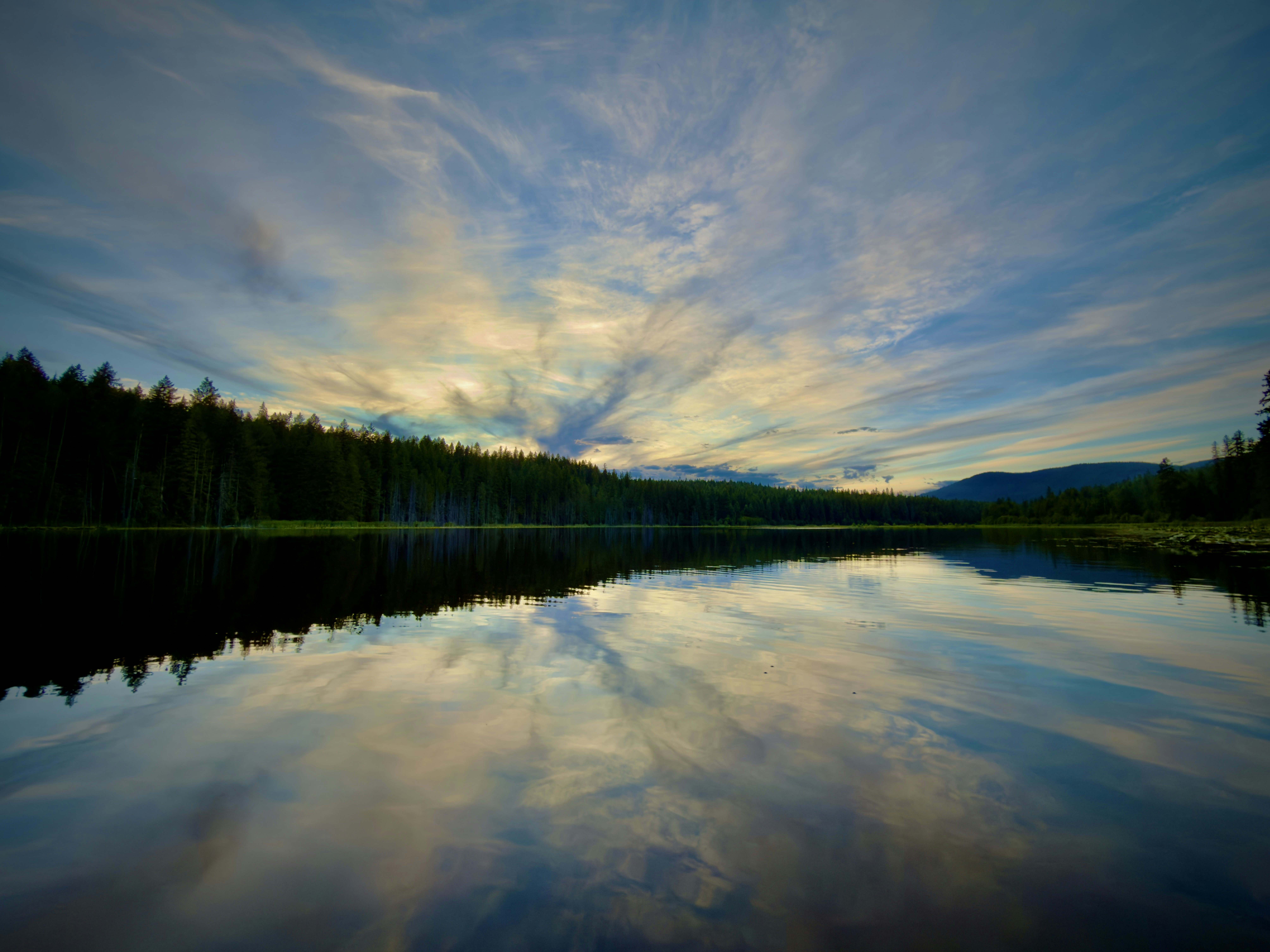 Serene lake scene with a forested shoreline and dramatic sky mirrored on the water, emphasizing the expansive horizon and tranquil ambience.