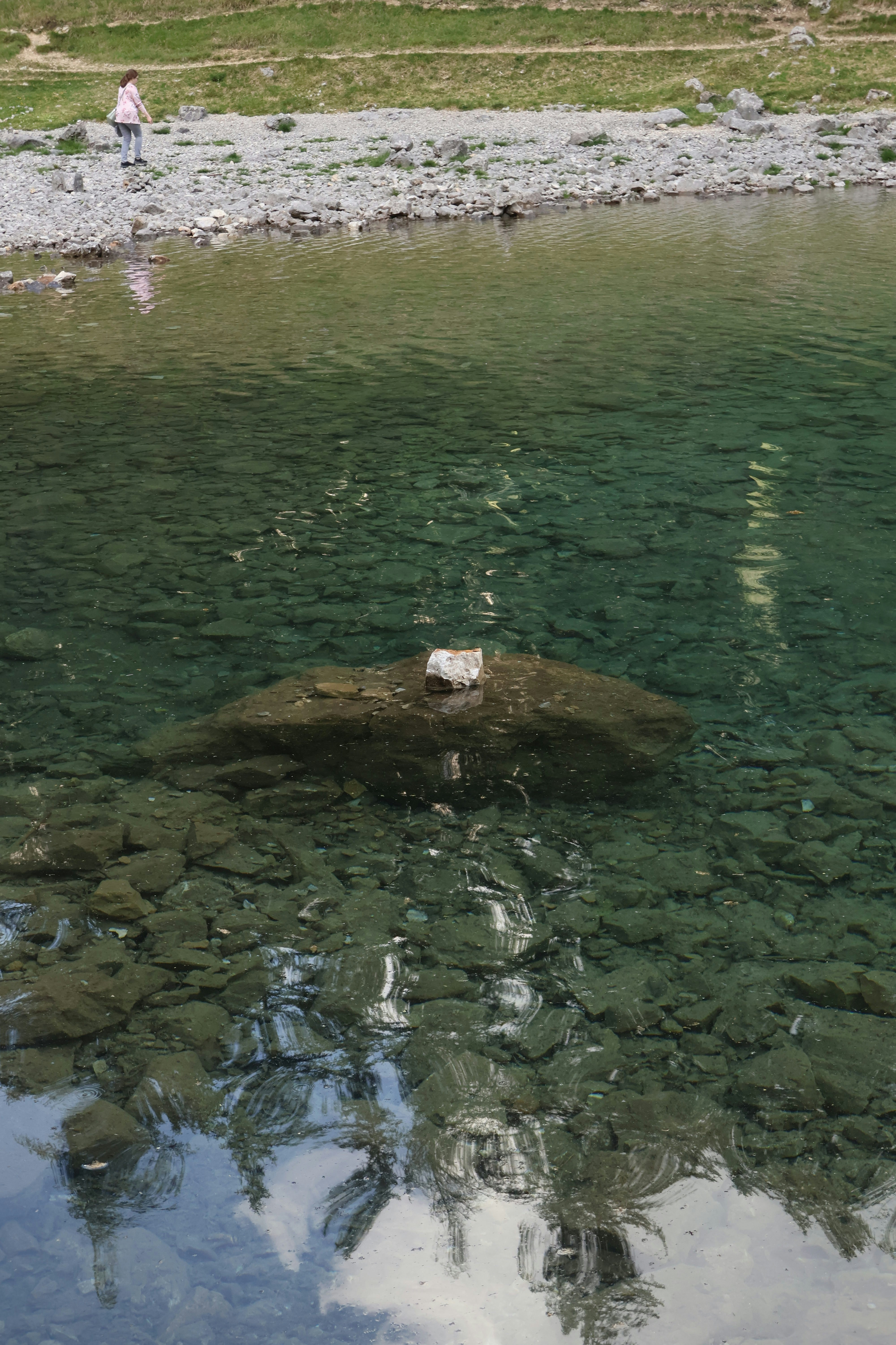 Clear river water over exposed rocks with a lone rock pinnacle near the center; a distant shoreline and a person on the pebbled bank provide scale. The scene emphasizes transparent shallows and natural textures.