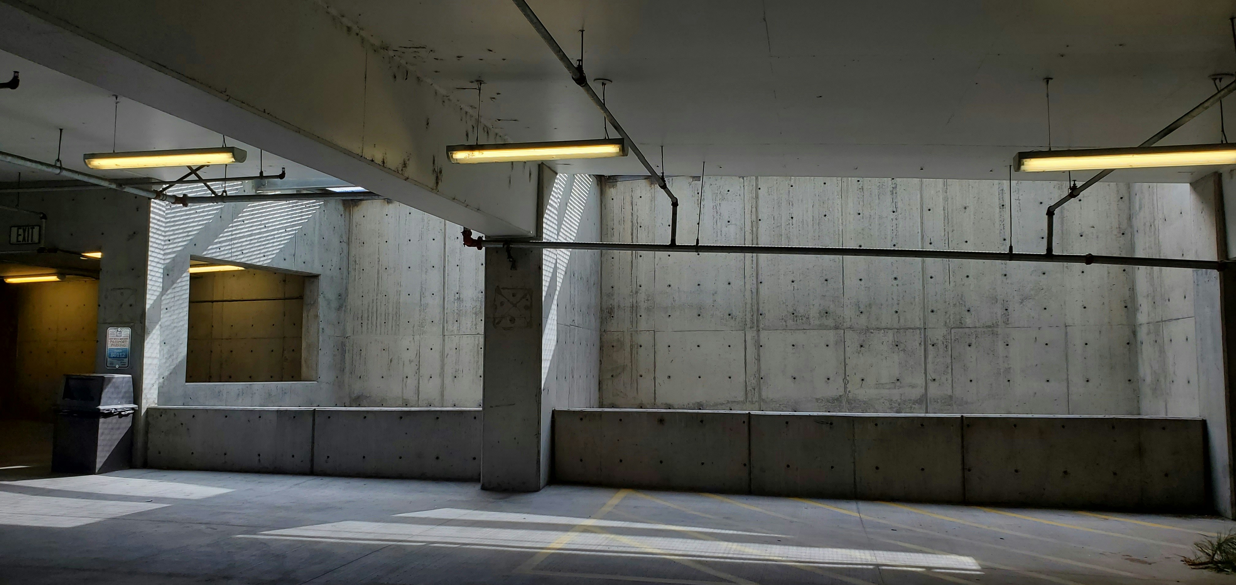 Wide-angle view of an empty concrete parking garage with overhead fluorescent lights and structural beams; minimal color, emphasizing harsh geometry and negative space. A minimal, architectural scene rather than a person or object.
