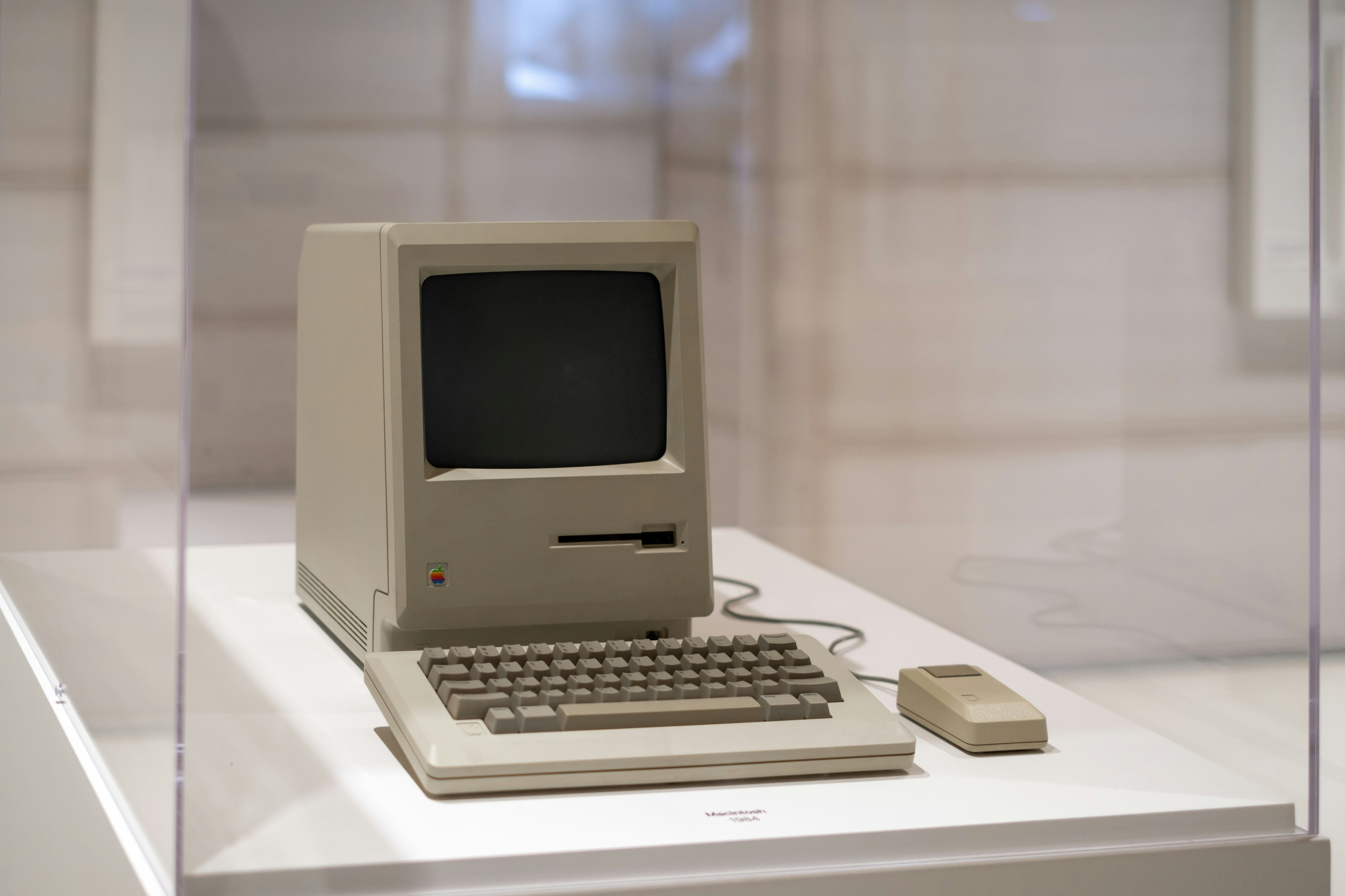 A vintage beige computer with a compact CRT monitor sits on a white pedestal in a museum display.