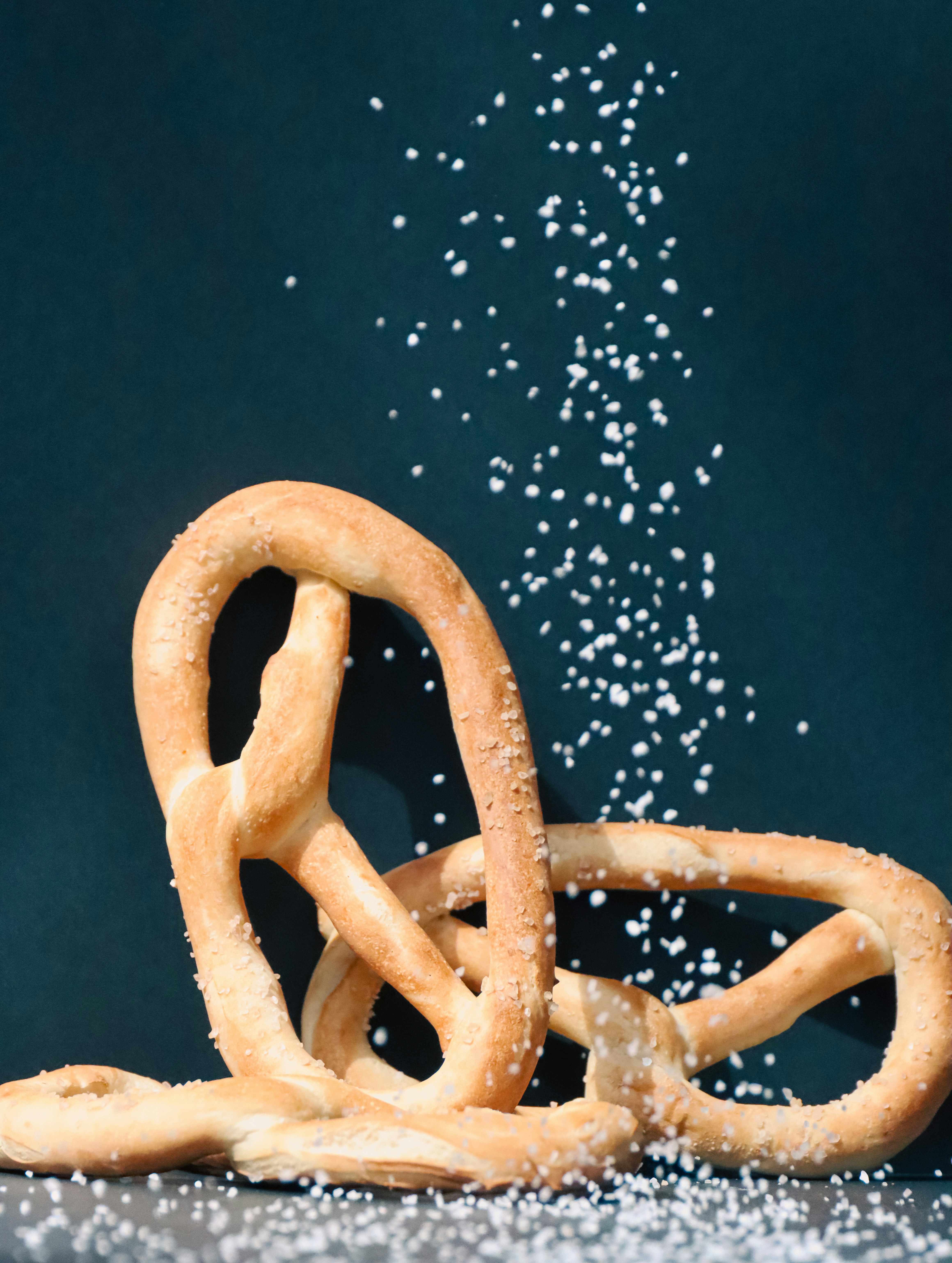 Golden pretzels stacked against a dark backdrop with salt crystals drifting around, capturing a simple still-life with a tactile, salty texture.