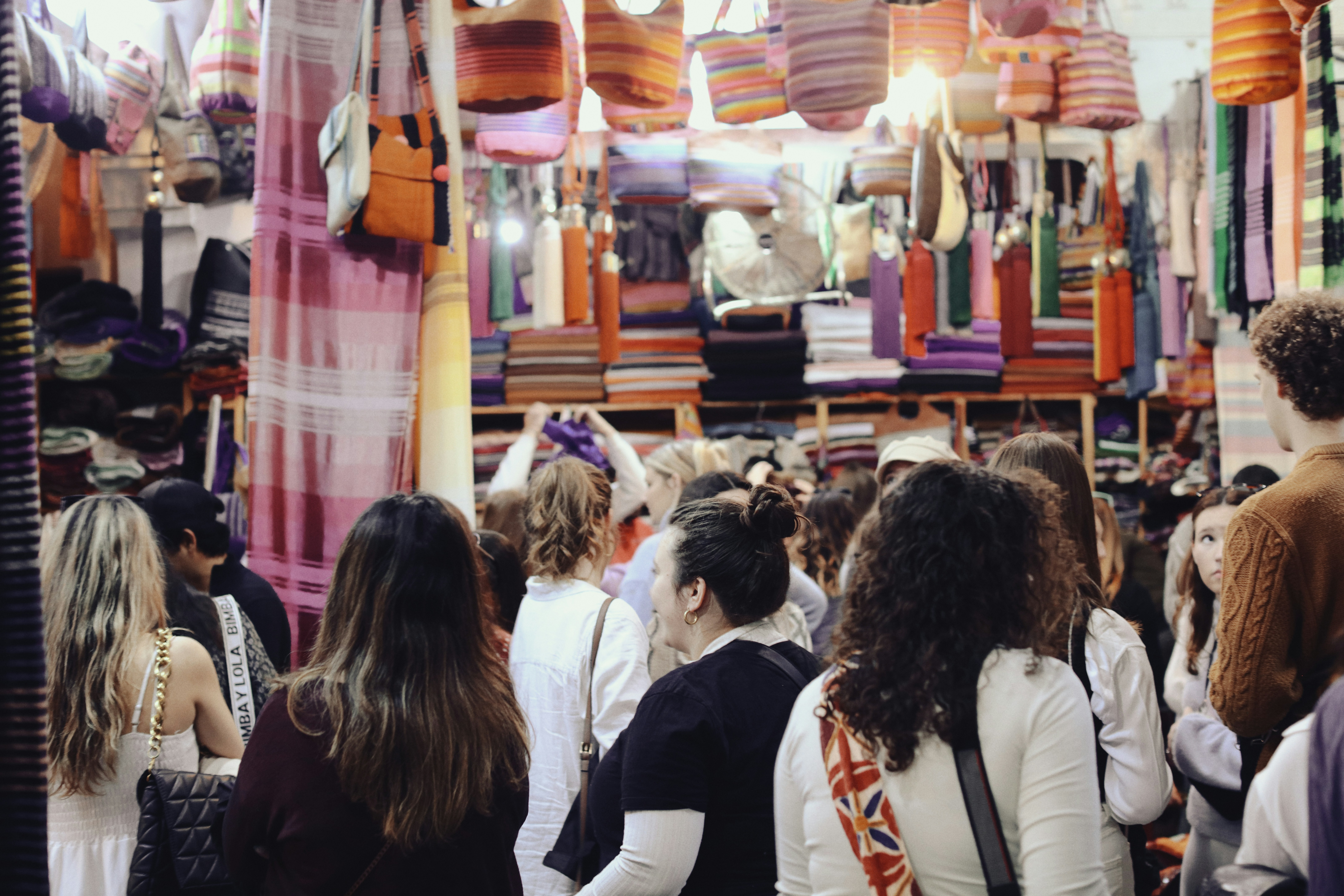Bustling market corridor filled with people amid hanging textiles and vibrant wares; a lively crowd scene showcasing shopfronts and colorful fabrics.