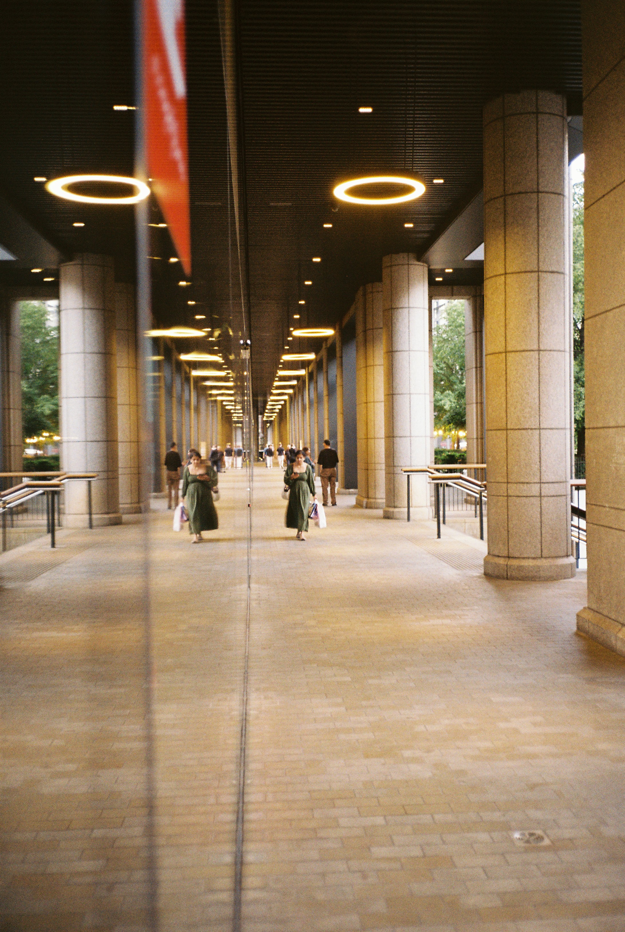 A woman in green walks a pillared colonnade lit by ring lights, doubled by a mirrored glass reflection.
