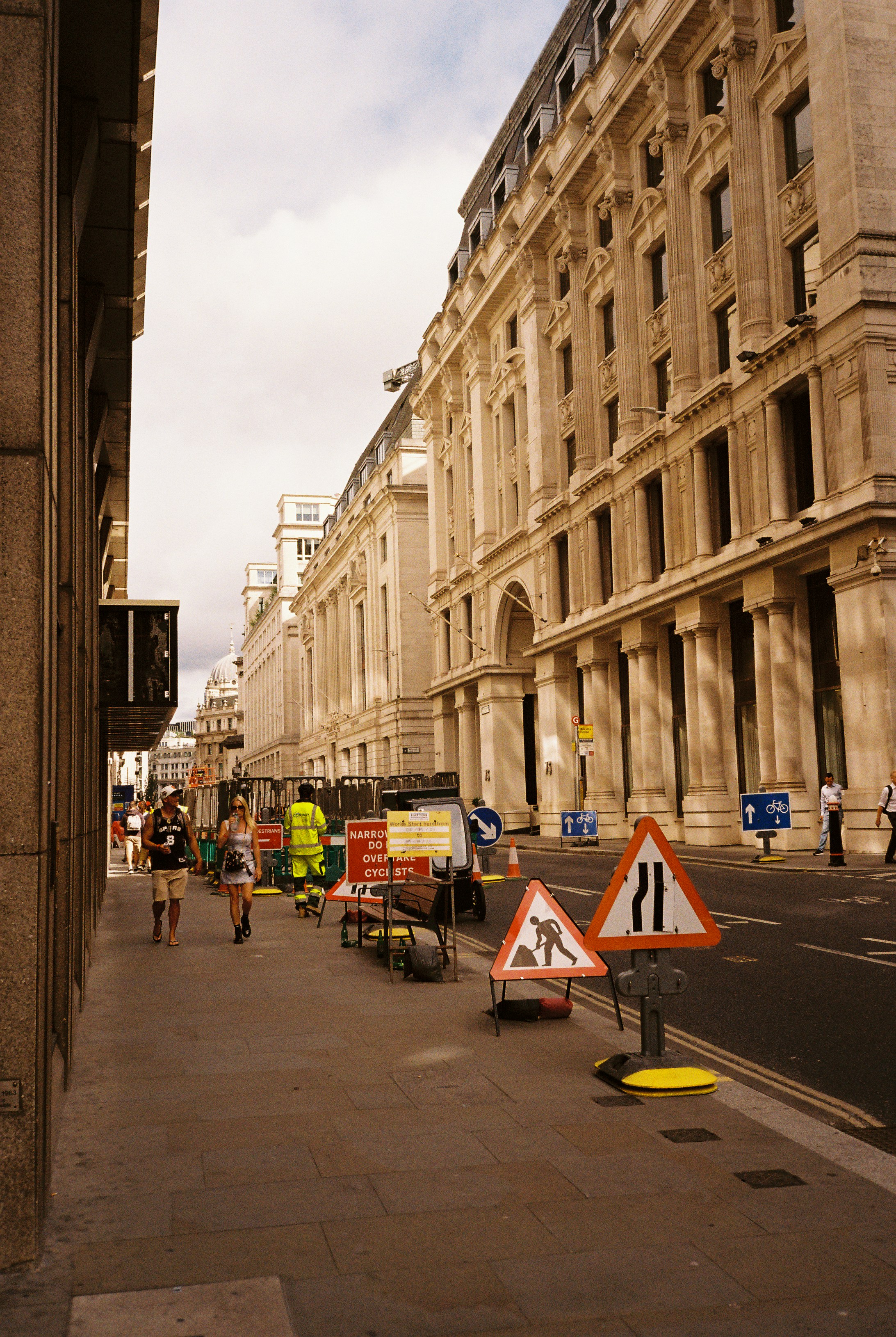 Pedestrian sidewalk with construction signs and workers beside a row of historic, ornate buildings along a city street.