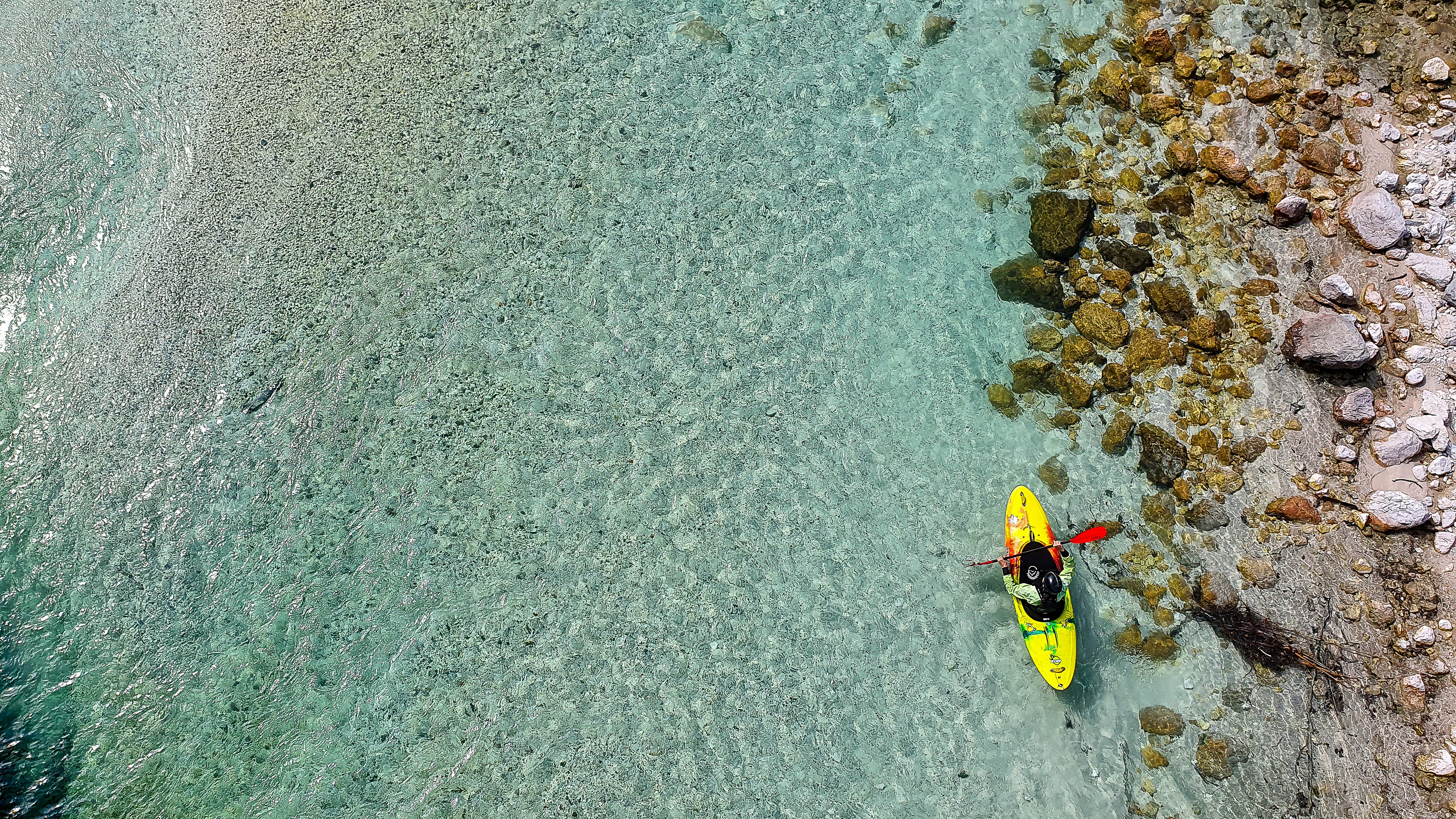 Bright yellow kayak with a lone paddler along a turquoise shoreline, rocks edging clear water, creating a calm coastal scene.