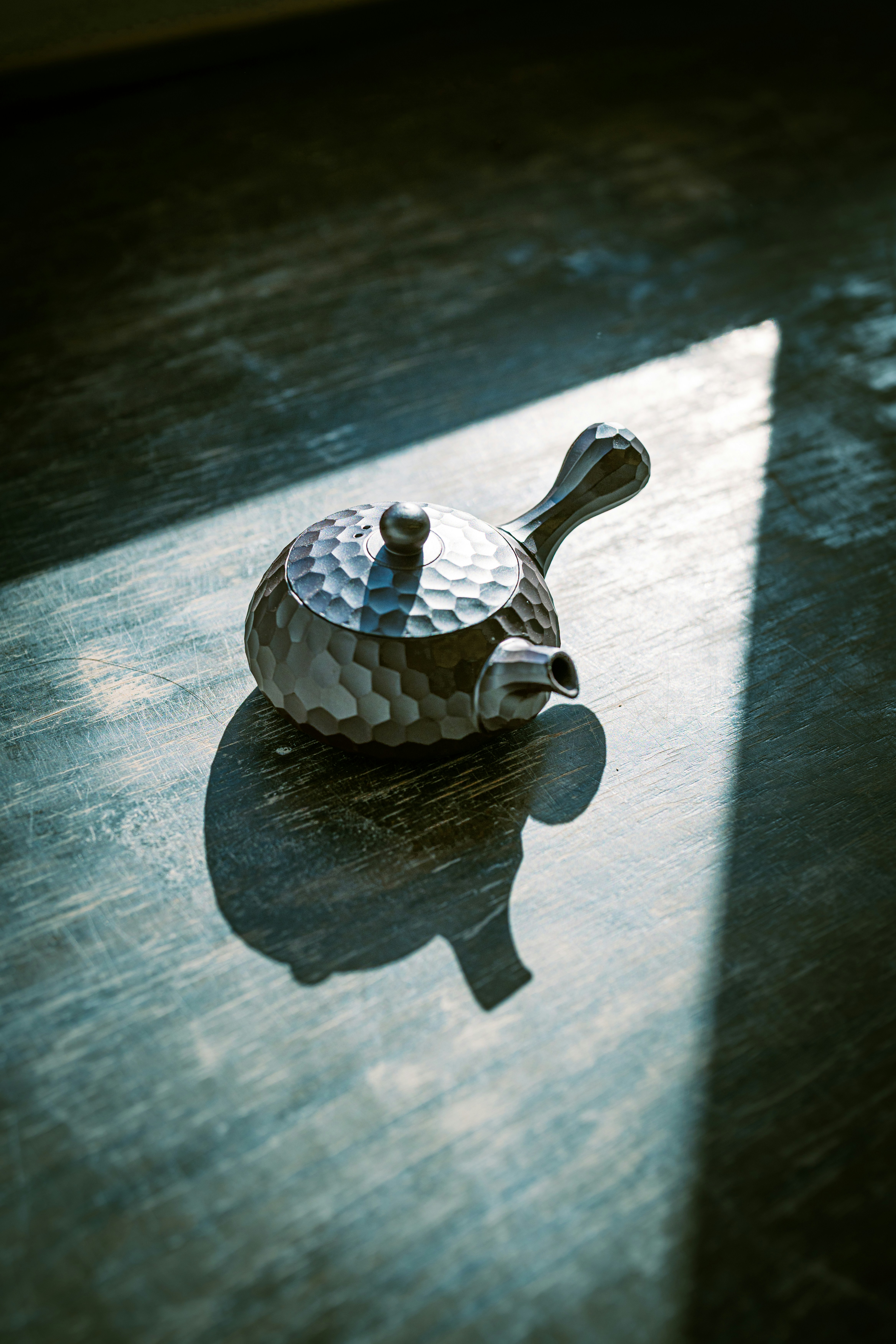 Steel-textured teapot sits on a dark wooden surface, illuminated by a diagonal beam of light casting a strong shadow; a shallow depth of field emphasizes the hexagonal pattern and sculpted handle. Photography print.