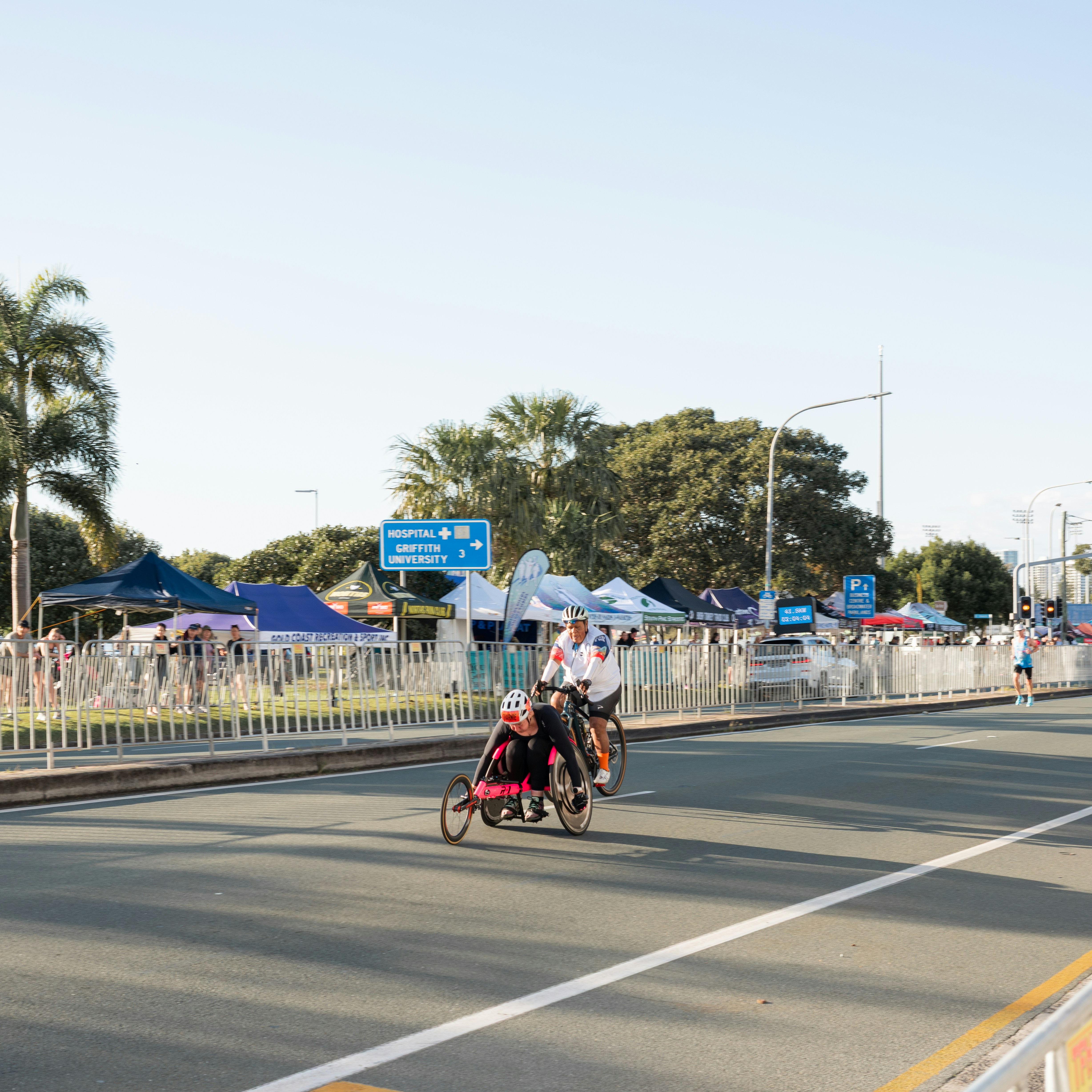 Wheelchair duo on a sunlit boulevard, race tents lining the curb