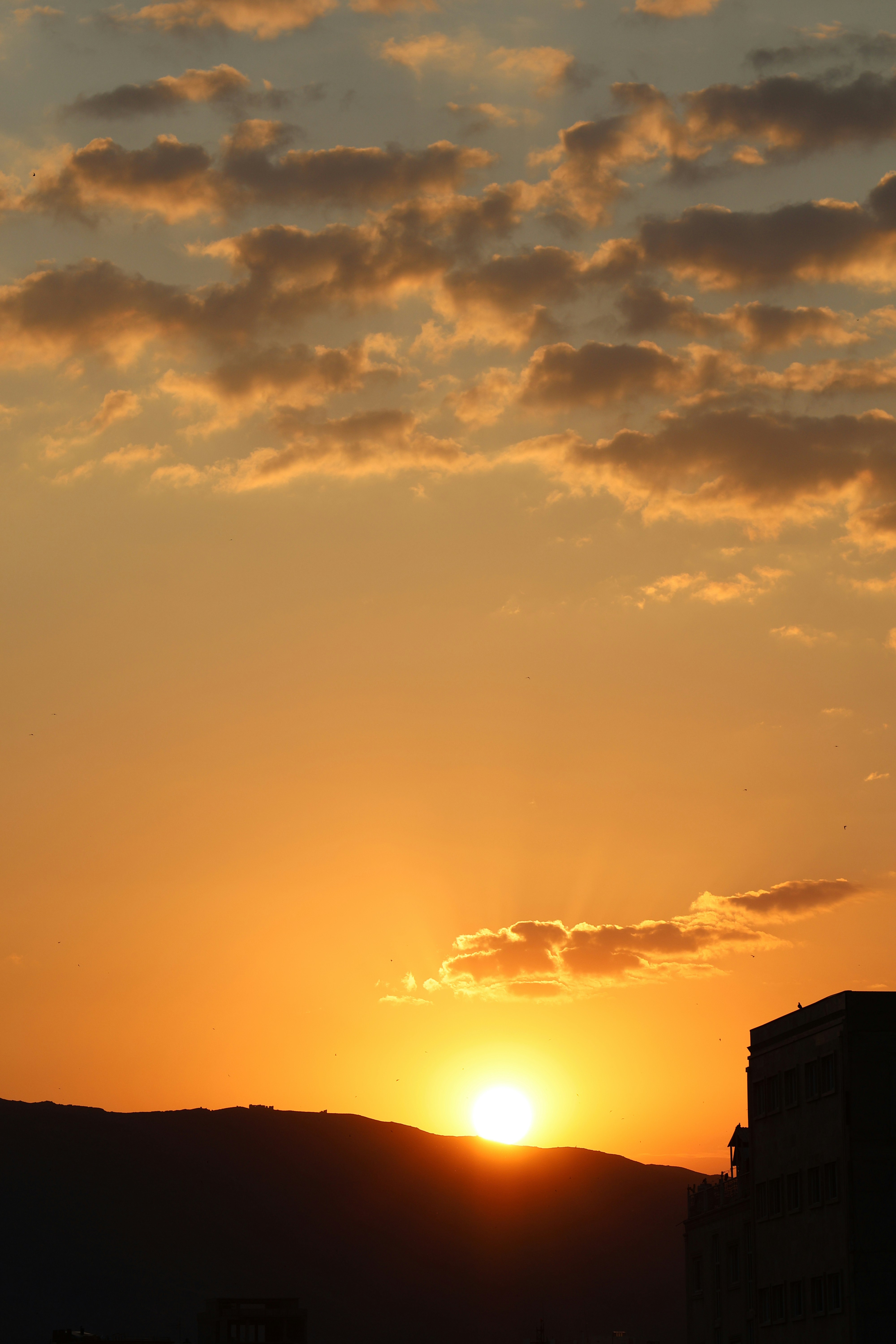 Golden sunset over a silhouetted cityscape with a glowing orb dipping behind distant hills, clouds catching warm hues.