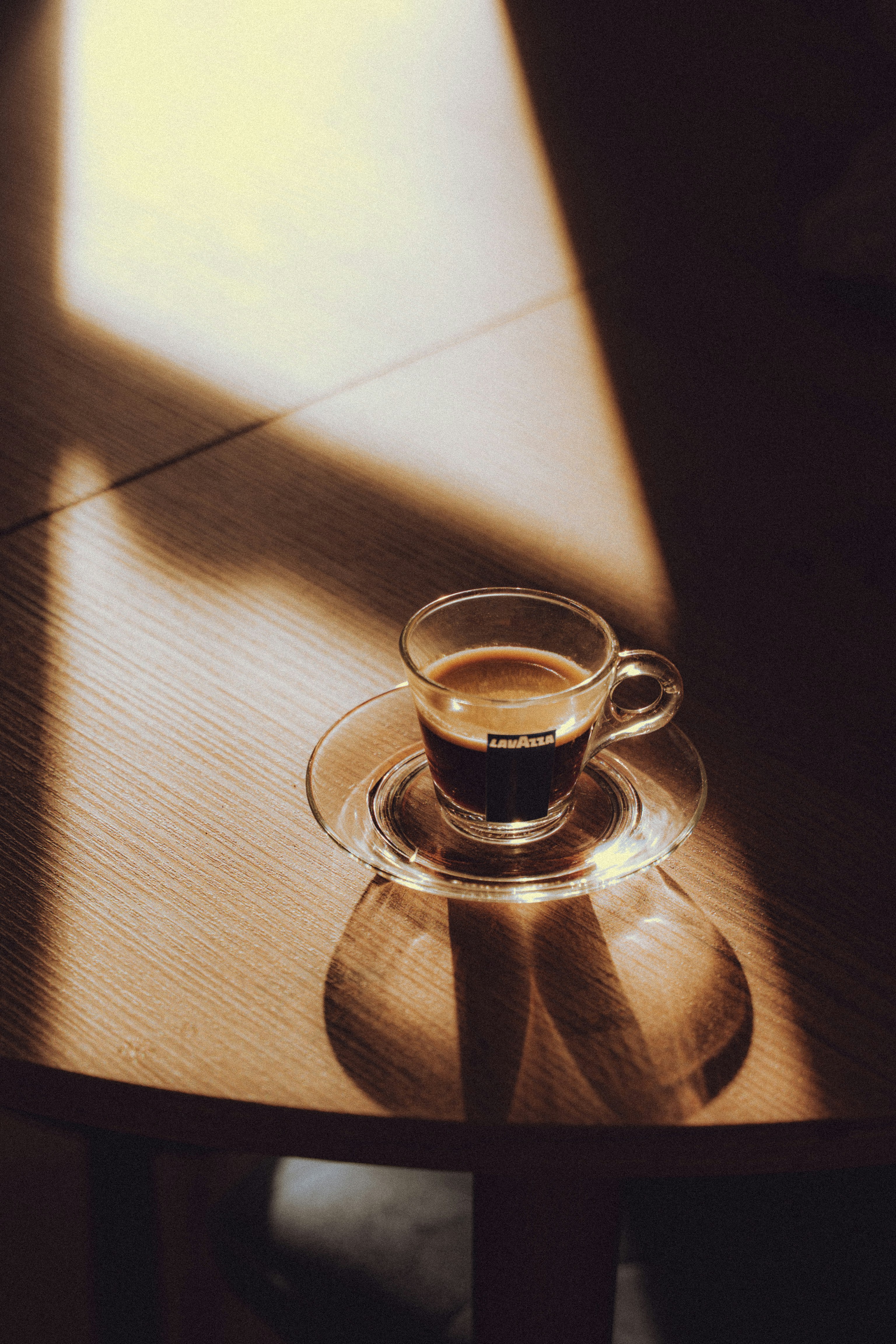 A small espresso cup sits on a glass saucer centered on a wooden table, bathed in warm, directional light that casts geometric shadows.