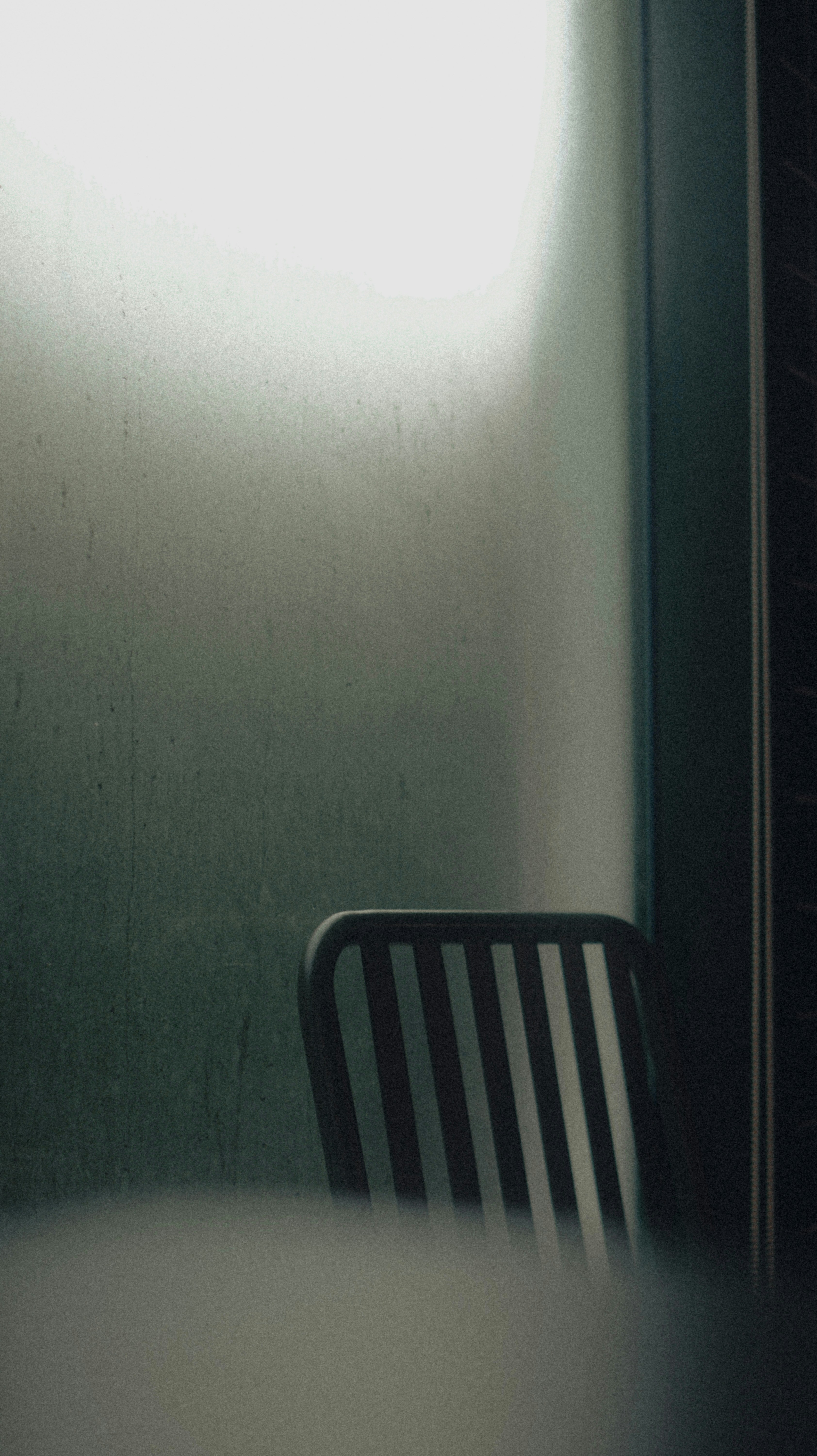 Low-contrast scene of a metal chair silhouette beside a foggy window; a soft, diffused light fills the upper left, emphasizing mood and minimalistic detail. Photograph has a documentary/industrial feel.