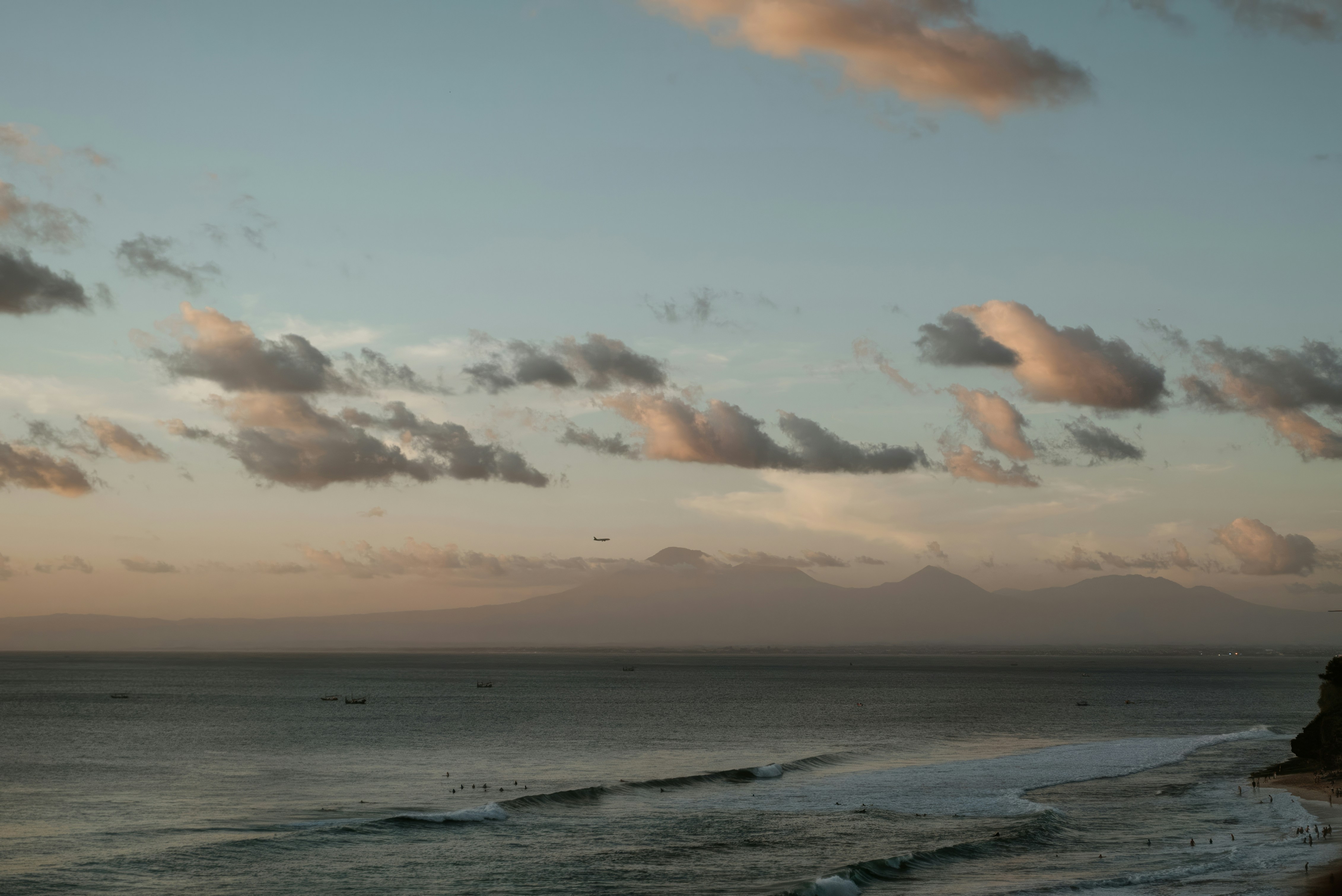 Soft coastal scene at sunset with distant misty landform and calm waves; a subtle silhouette on the right hints at a shoreline feature.
