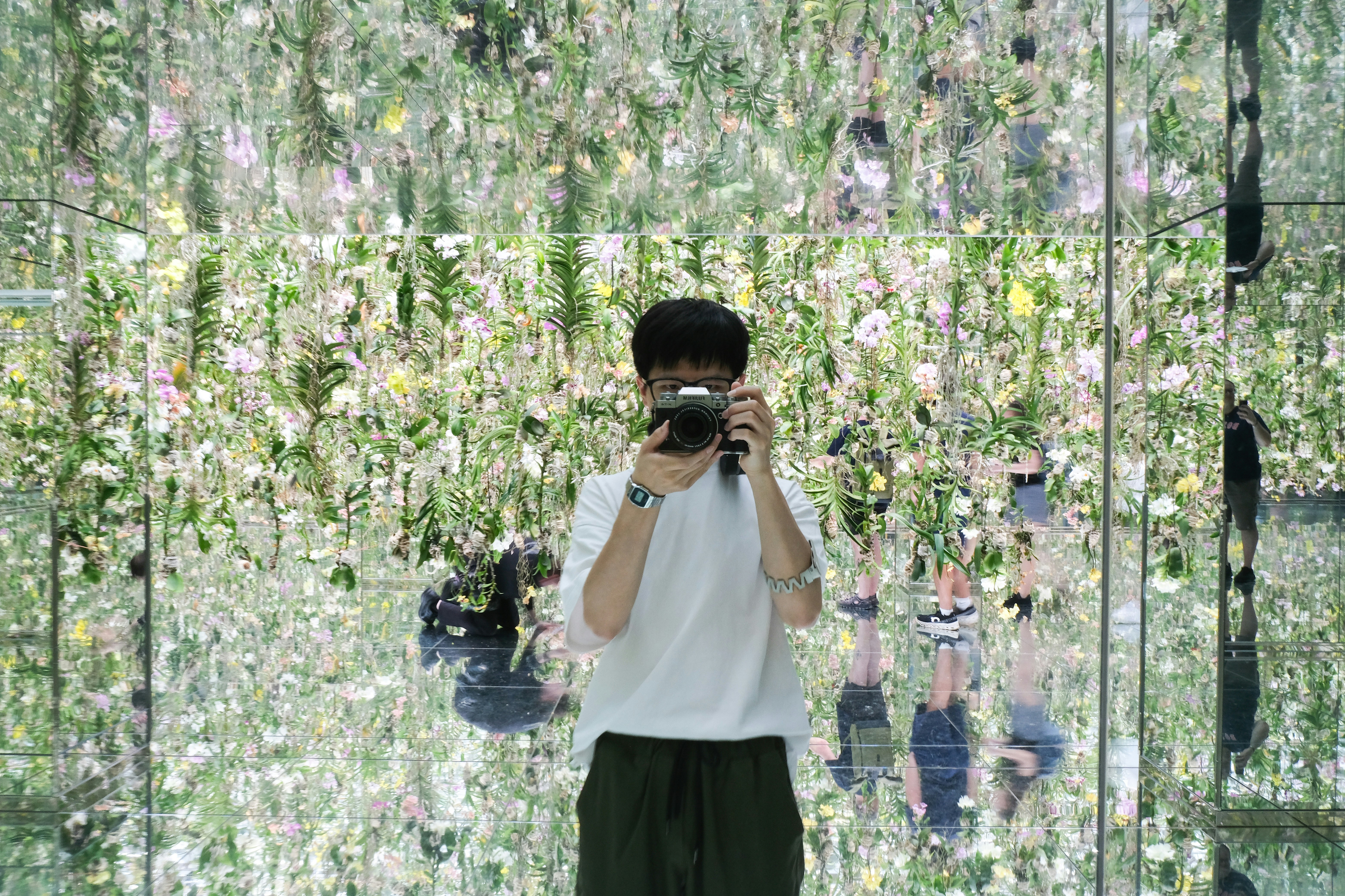 A person with a camera stands center-stage in a mirrored floral installation, capturing their reflection amidst hanging blossoms. This is a photographic scene inside a gallery-like space with a lush, mirrored backdrop.