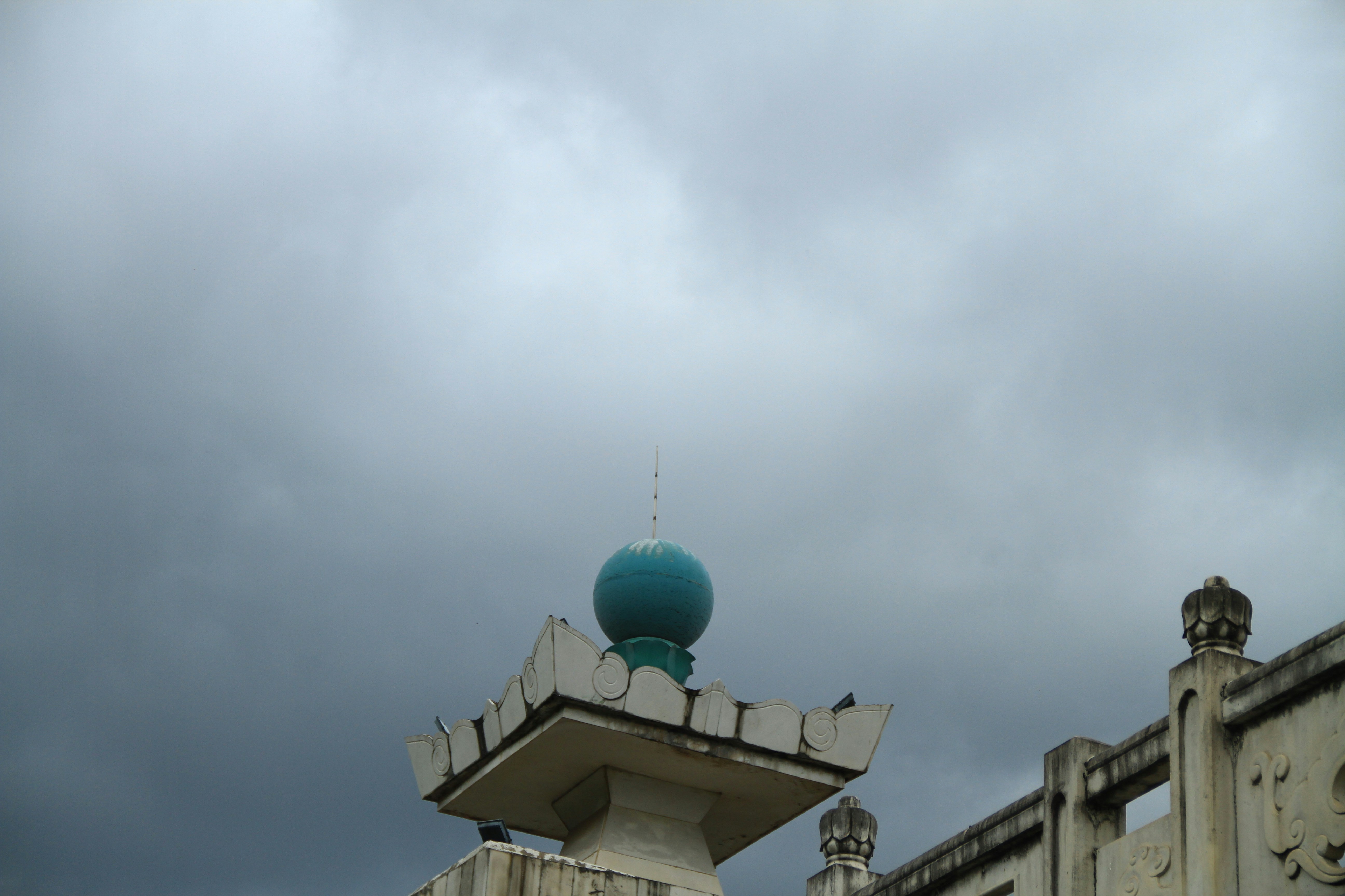 A turquoise dome sits atop a decorative structure against a moody overcast sky; architectural details and railing are visible in the foreground. This is a photographed architectural detail from a temple or monument.