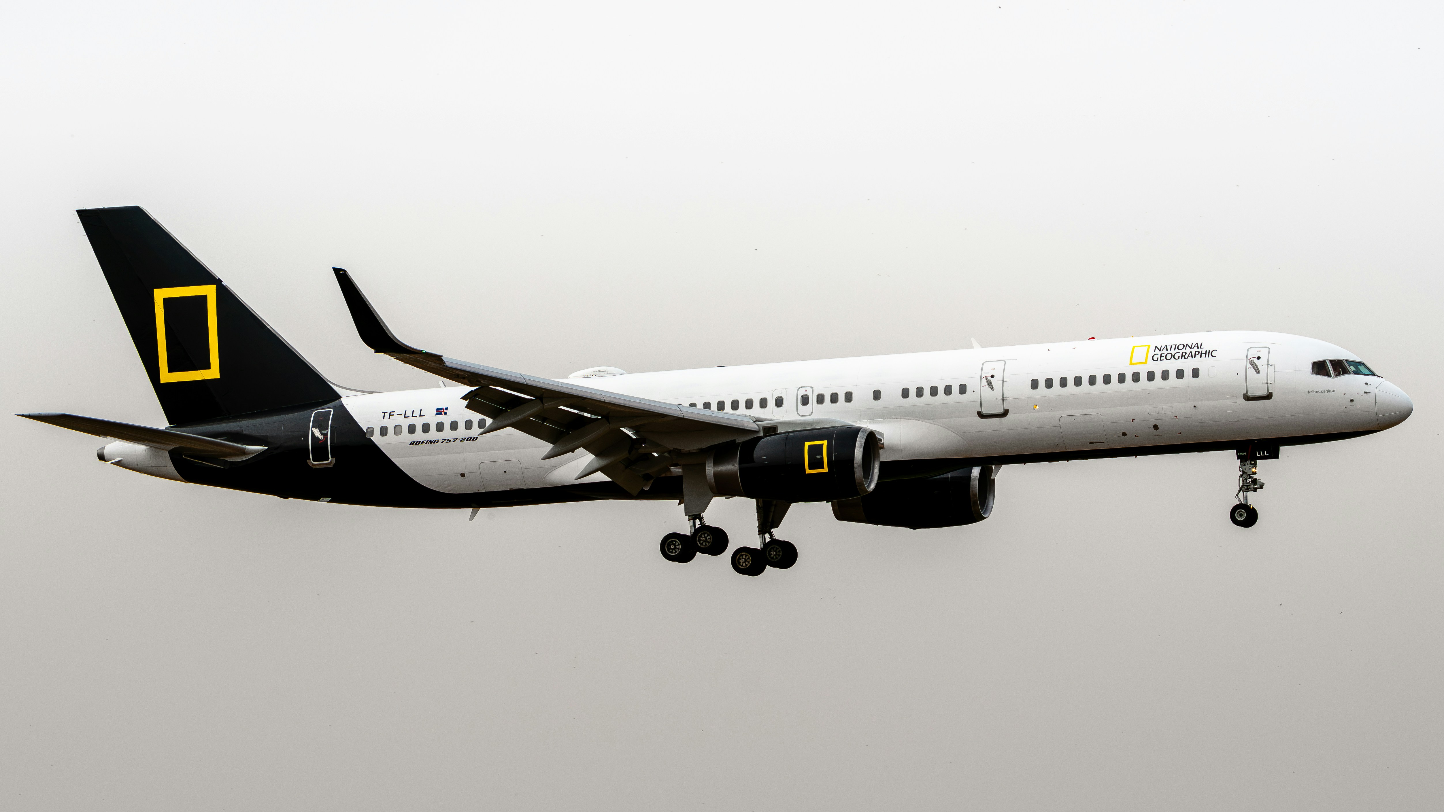 A National Geographic-branded passenger jet descends against a pale sky, its livery and tailmark clearly visible as landing gear deploys. The image emphasizes aircraft design and aviation branding in a minimalist backdrop.