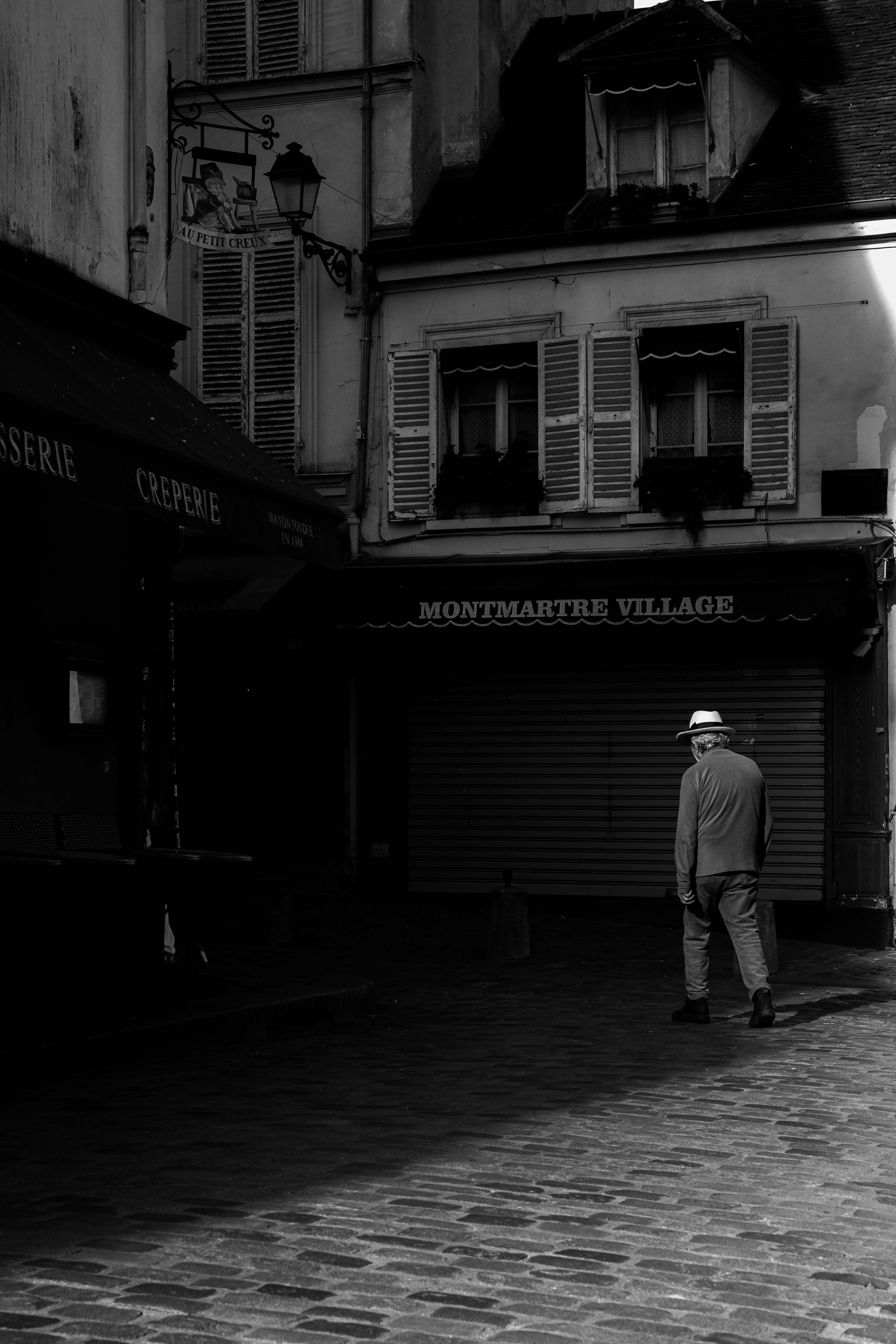 Moonlit street scene with a solitary man in a hat walking away from the camera; weathered buildings and a shuttered storefront frame the quiet alley. A minimalist, monochrome study of solitude and urban texture.