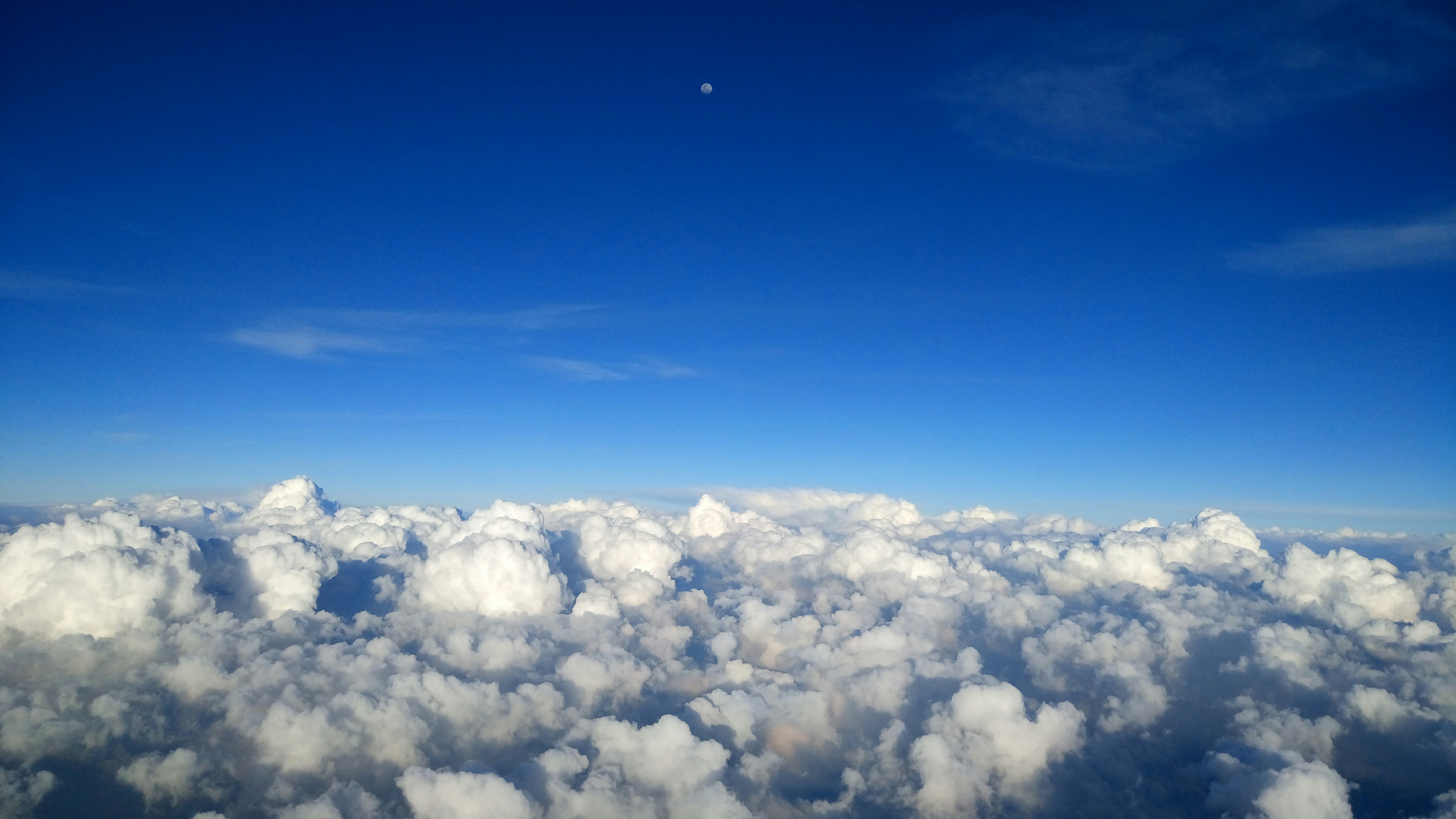 High altitude sky over a dense sea of clouds with a distant moon visible, capturing a wide, airy horizon. The scene emphasizes vast atmosphere and calm serenity.