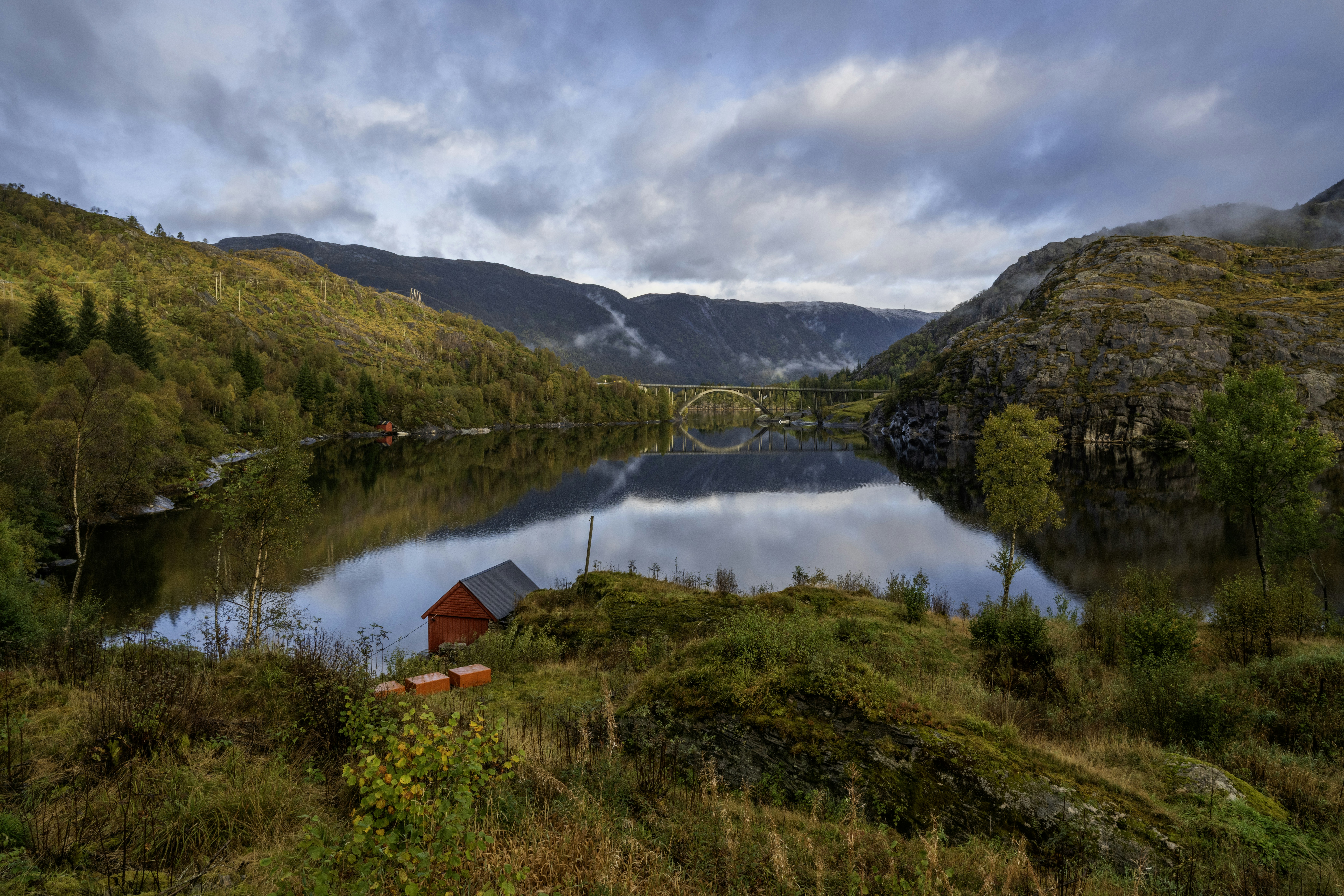 Red boathouse at the edge of a glassy fjord under a dawn-kissed ridge