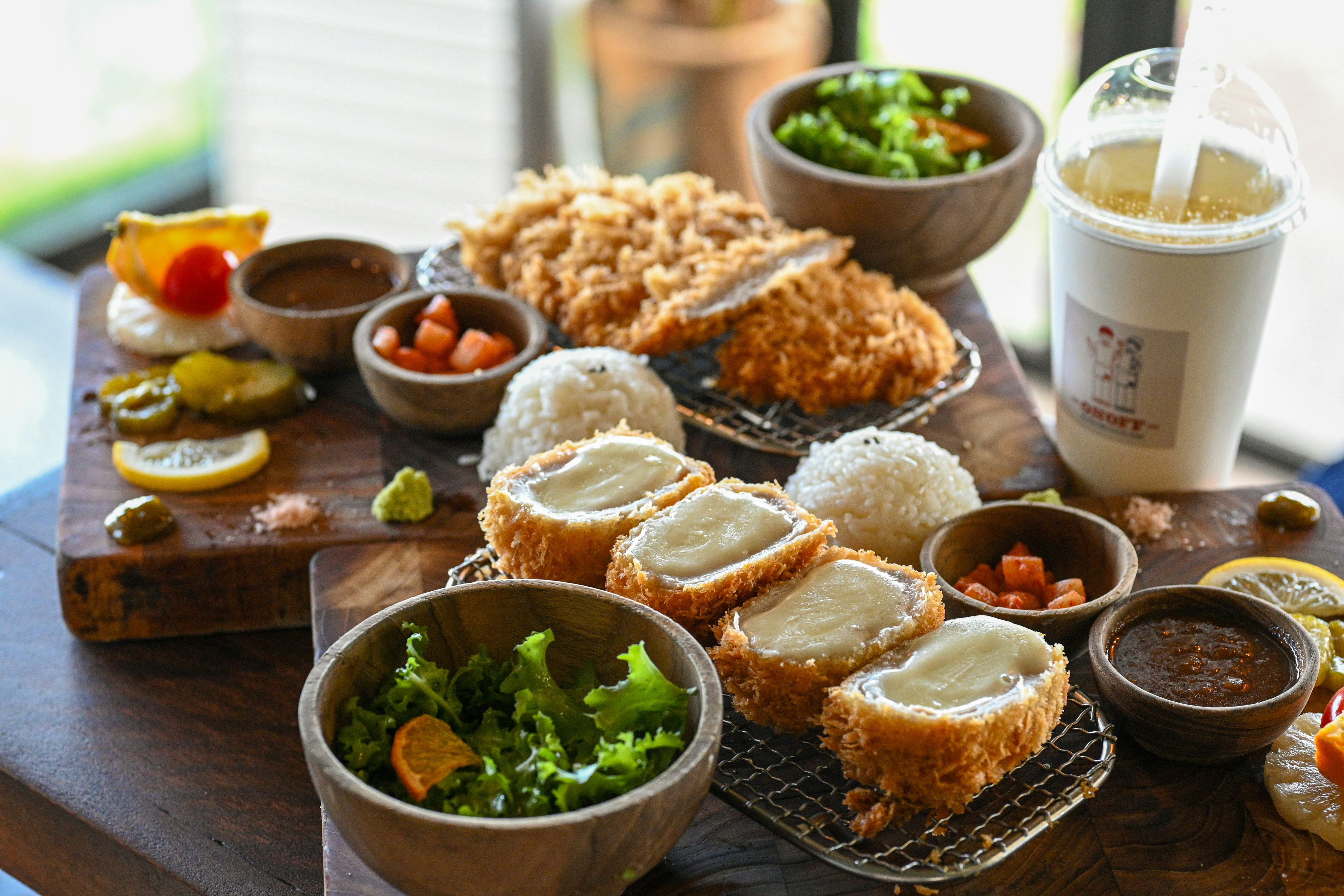 Meal spread featuring breaded cutlets with melted cheese, small bowls of condiments, greens, and a creamy beverage on a wooden board.