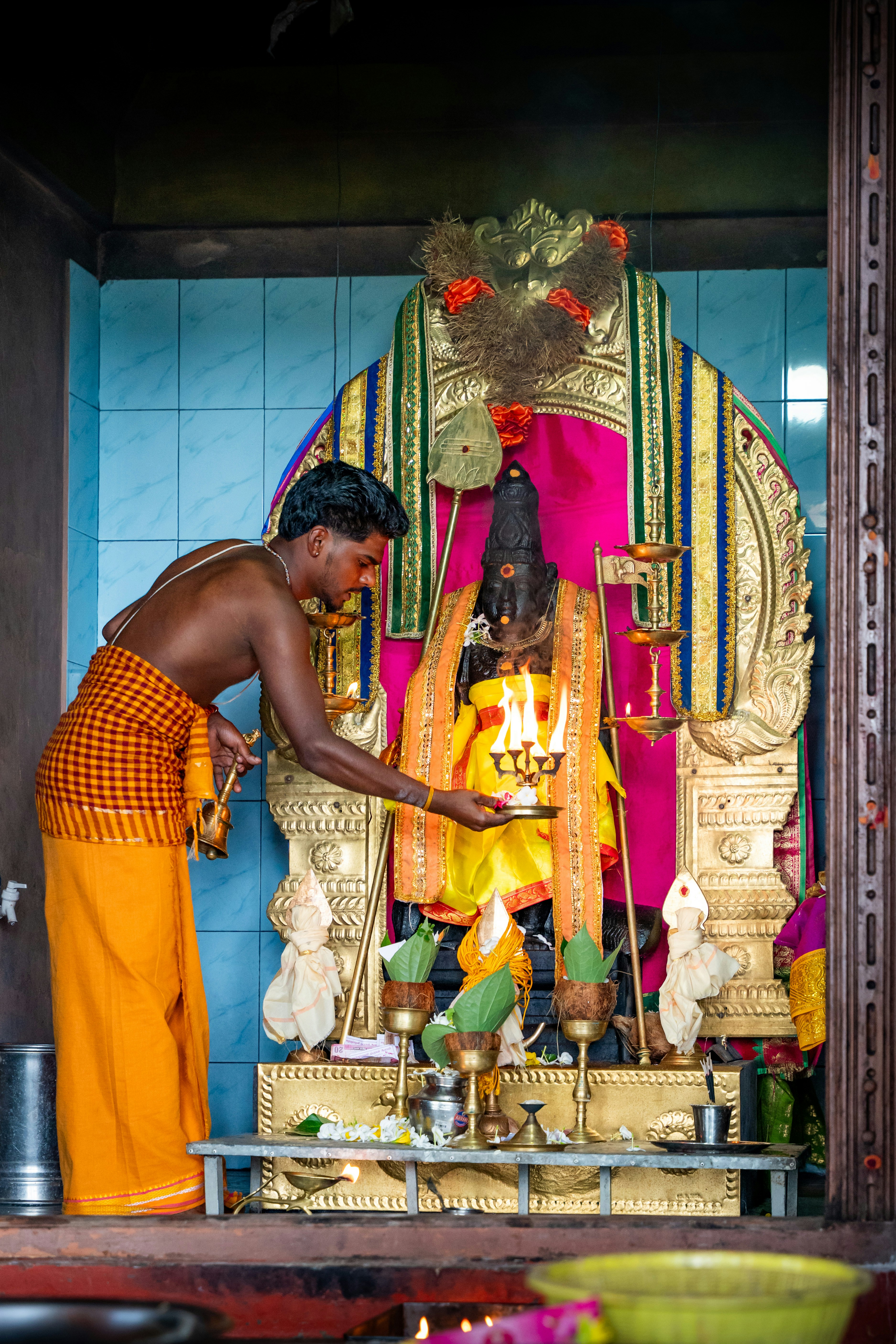 A devotee in bright saffron garments tends a lit oil lamp before a gilded Hindu statue, within a temple alcove adorned with flowers and ceremonial items.