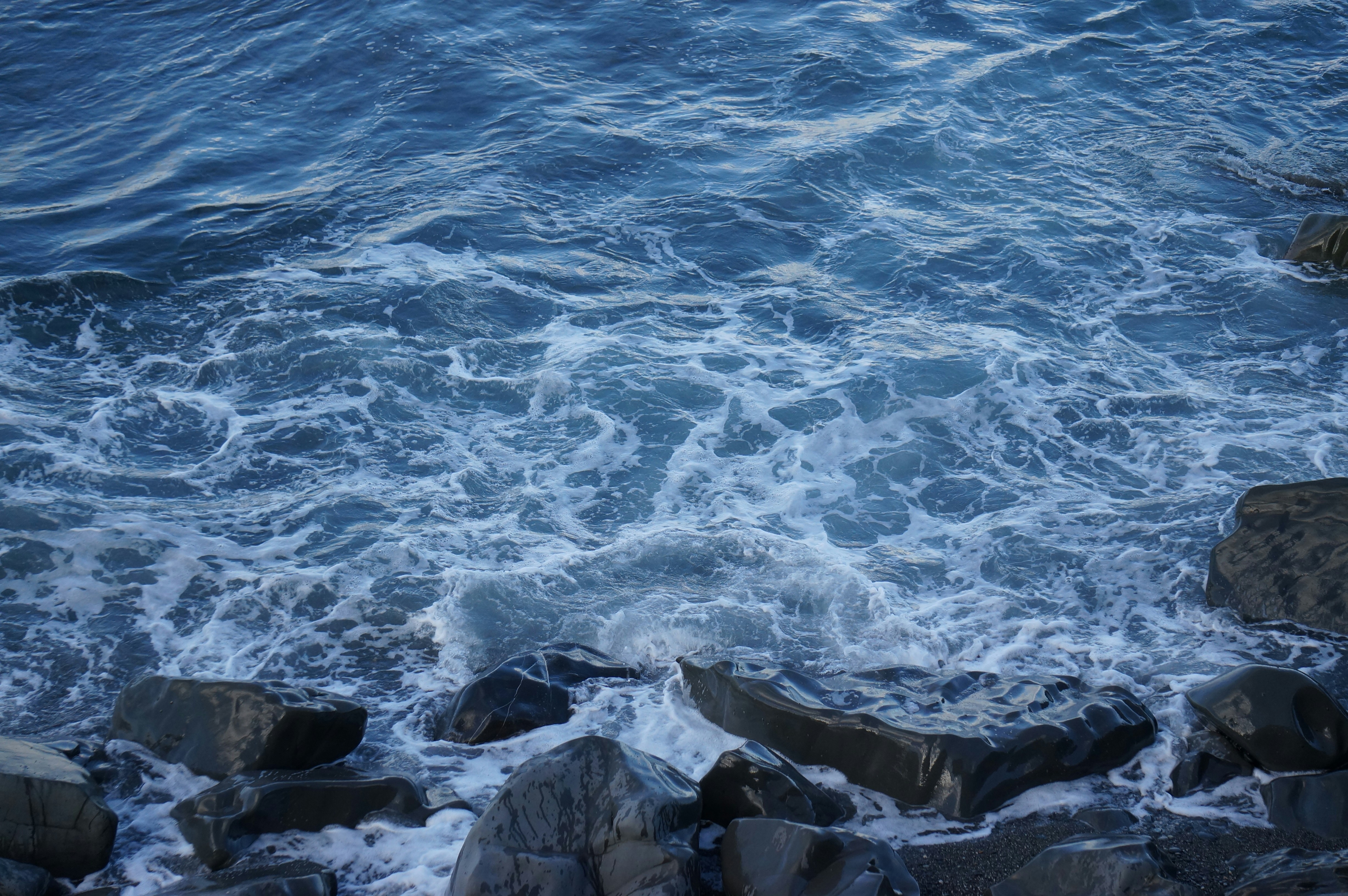 Coastal scene showing foamy waves colliding with dark rocks along a rugged shoreline. The image captures the textures of water and stone in a cool, moody palette.