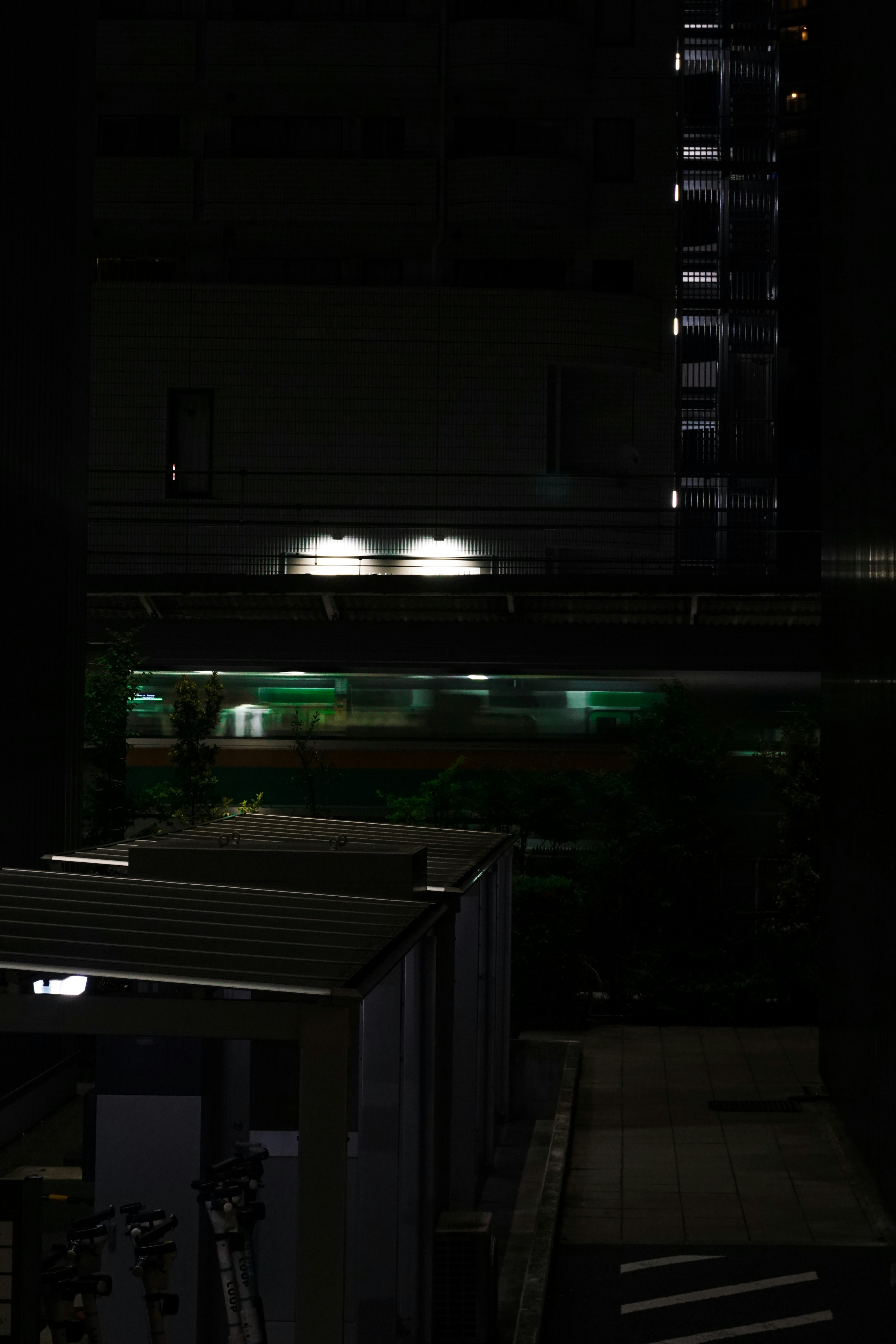 Nighttime urban scene with a passing green-lit train blurred across an elevated track; foreground features architectural canopies and walkways, hinting at a quiet, industrial courtyard.