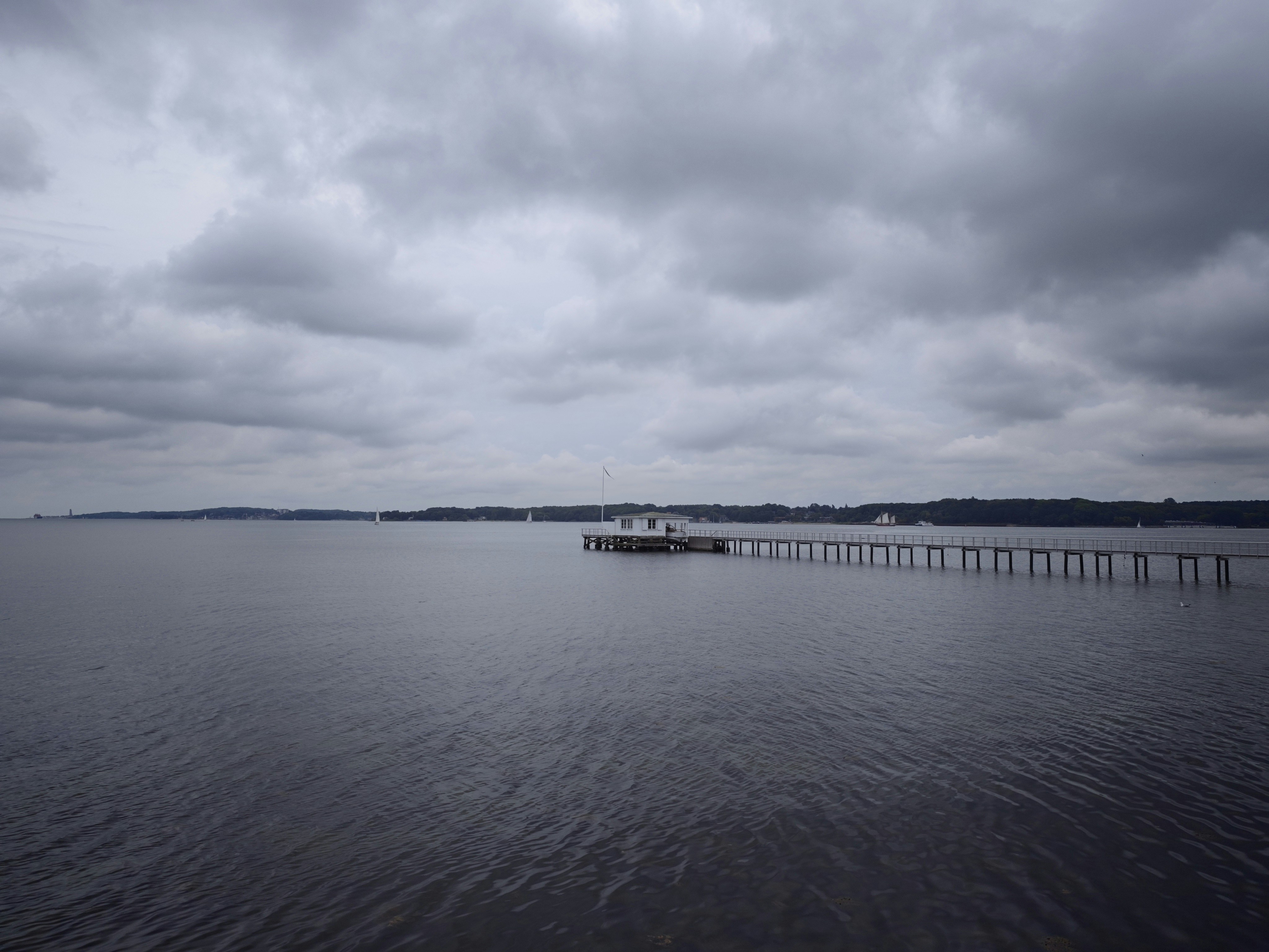 Long pier stretches into calm water under moody skies, with distant shore on the horizon and gentle ripples in the foreground. A subdued coastal scene devoid of people or boats.