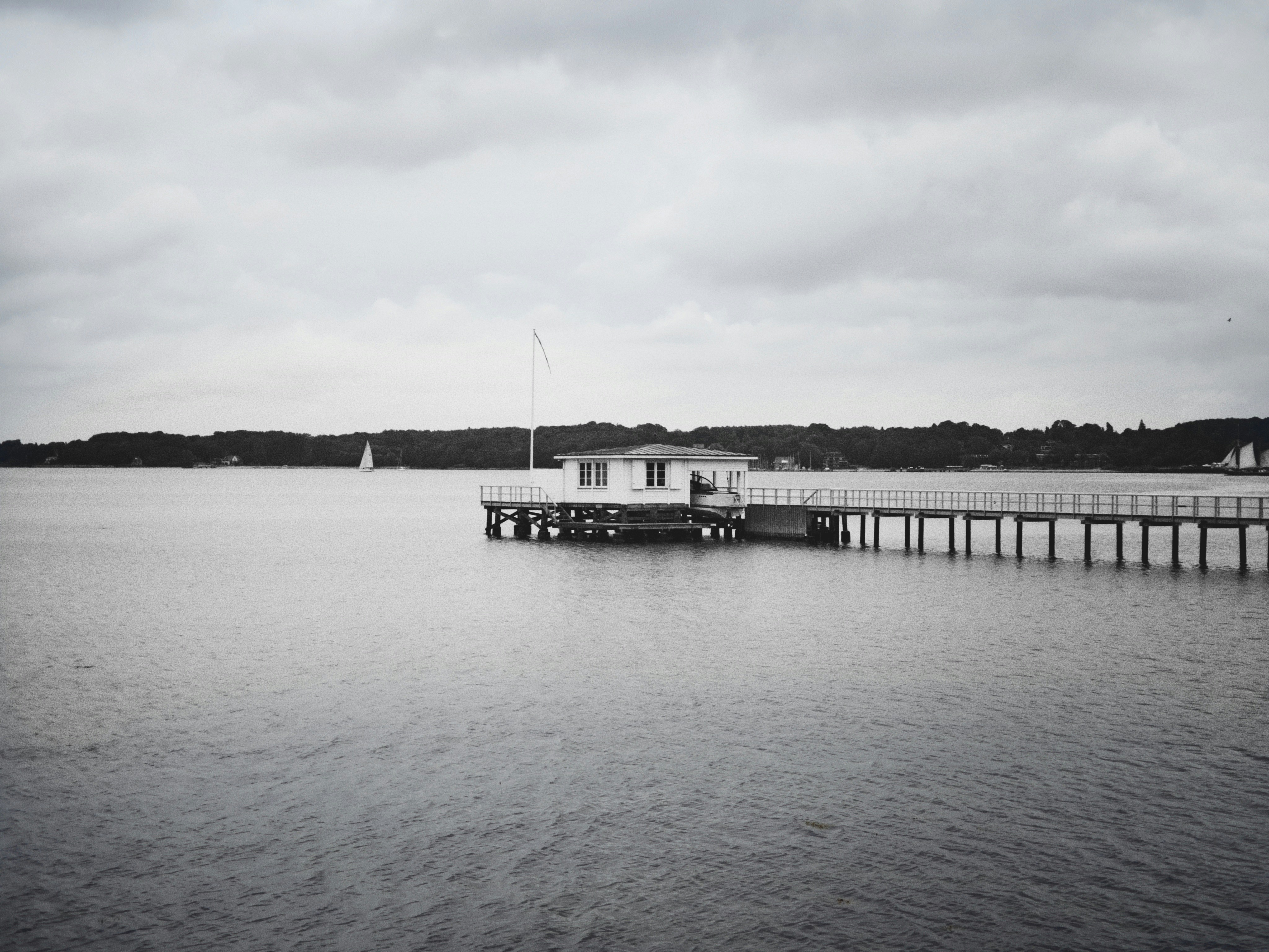 A tranquil grayscale scene of a small house on a pier extending into calm water, with a distant shoreline and sailboats on the horizon.