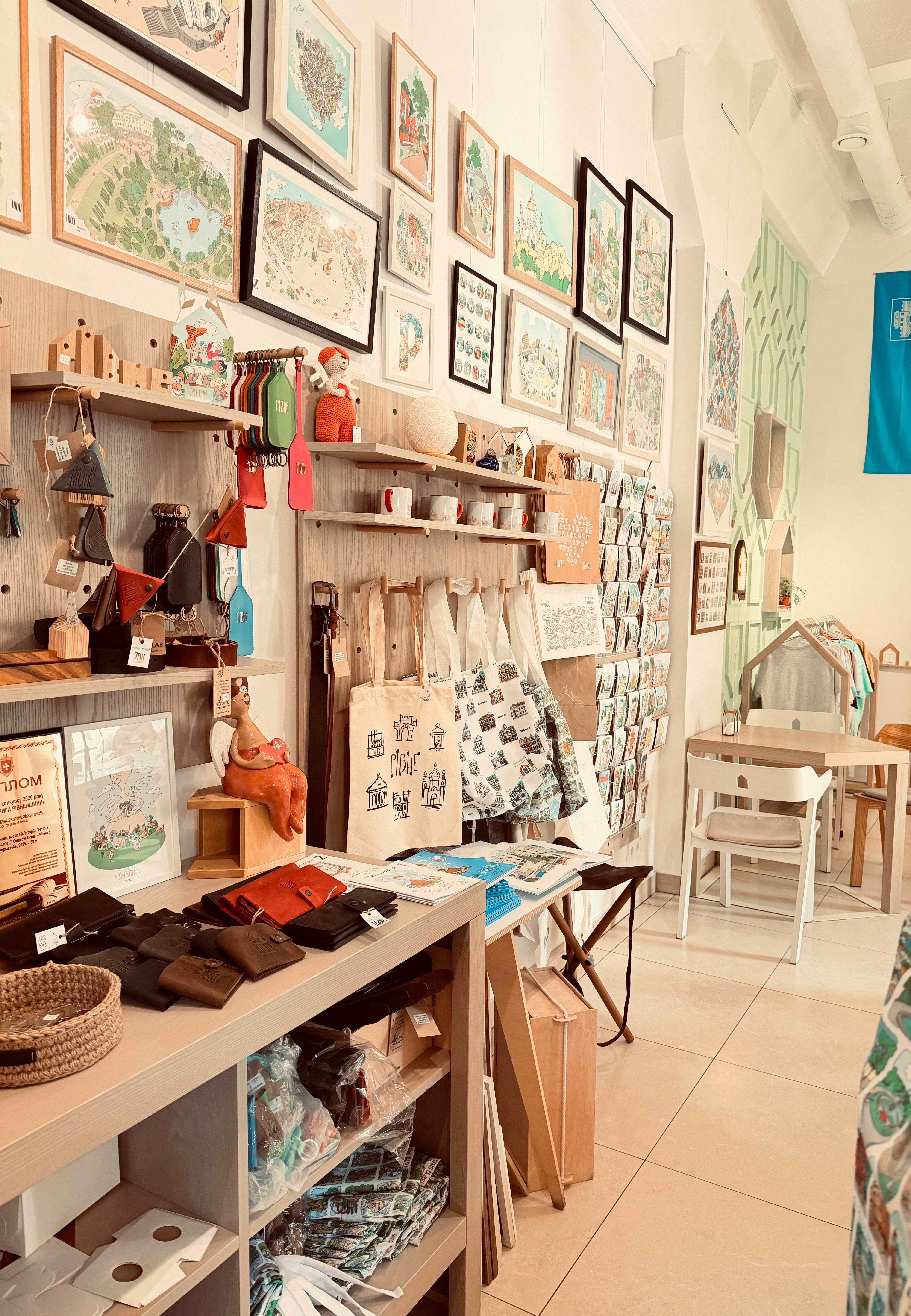 A bright interior shop with walls lined by framed maps, shelves of handmade goods, and tables displaying textiles and accessories. This image captures a cozy storefront or studio space featuring assorted crafts.