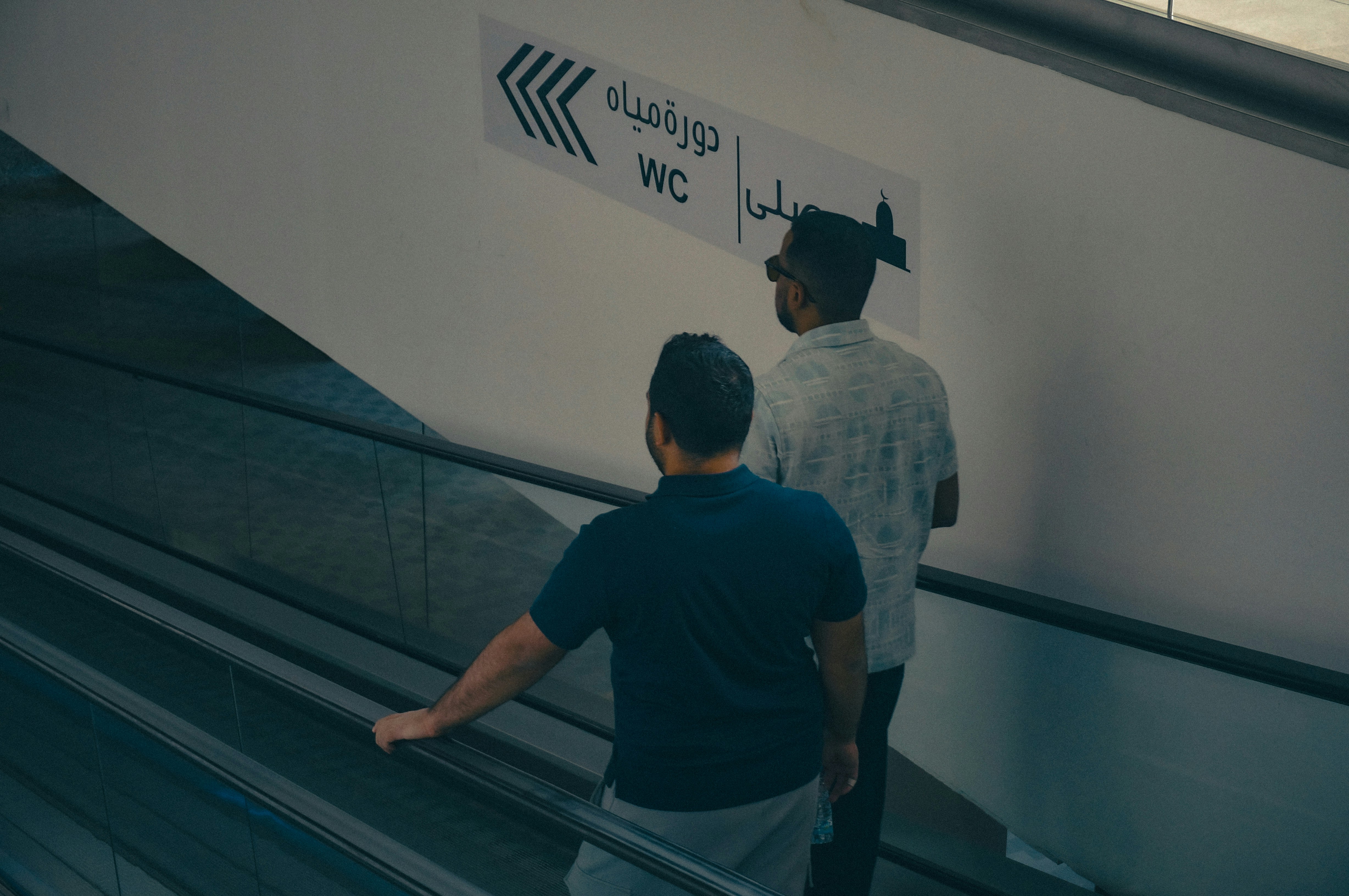 Escalator Ascent: Two Travelers Approach a Sign toward the WC
