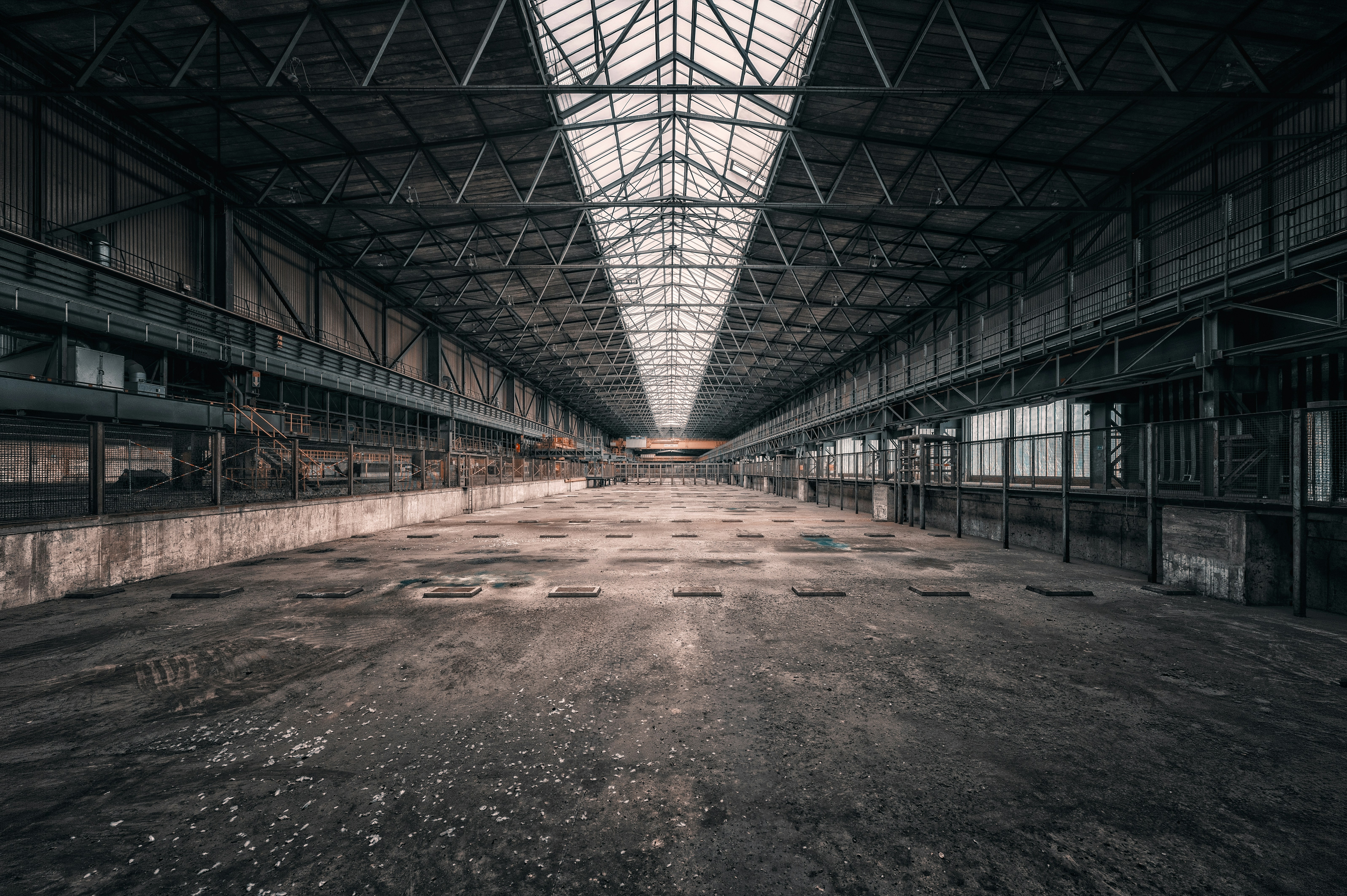 Deserted industrial interior with a long, converging aisle under a skylit roof, emphasizing symmetry and texture. A stark, monochrome scene highlighting worn concrete and steel framework.