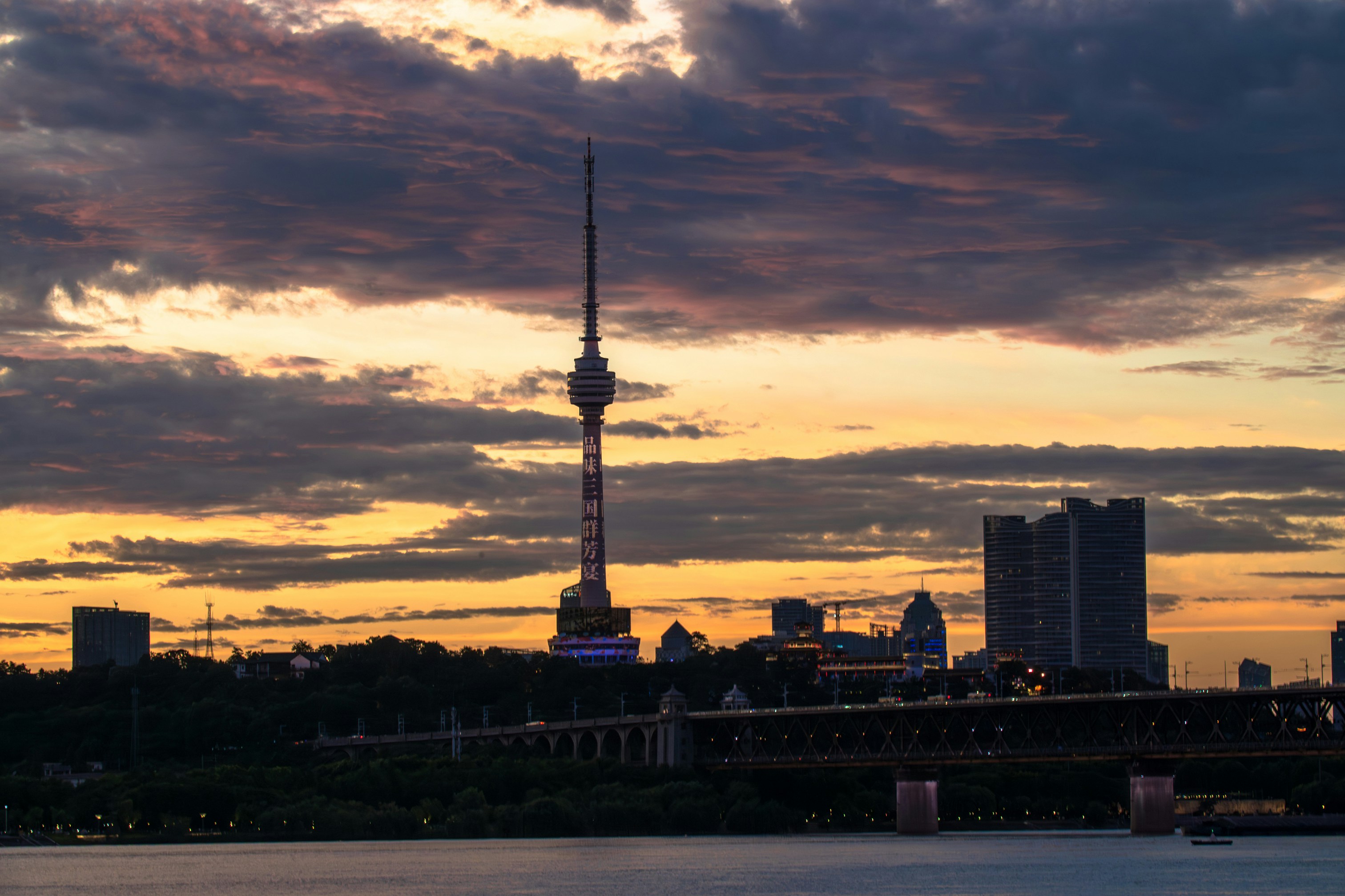 Vivid sunset over a city skyline with a tall tower and bridges spanning a calm river.