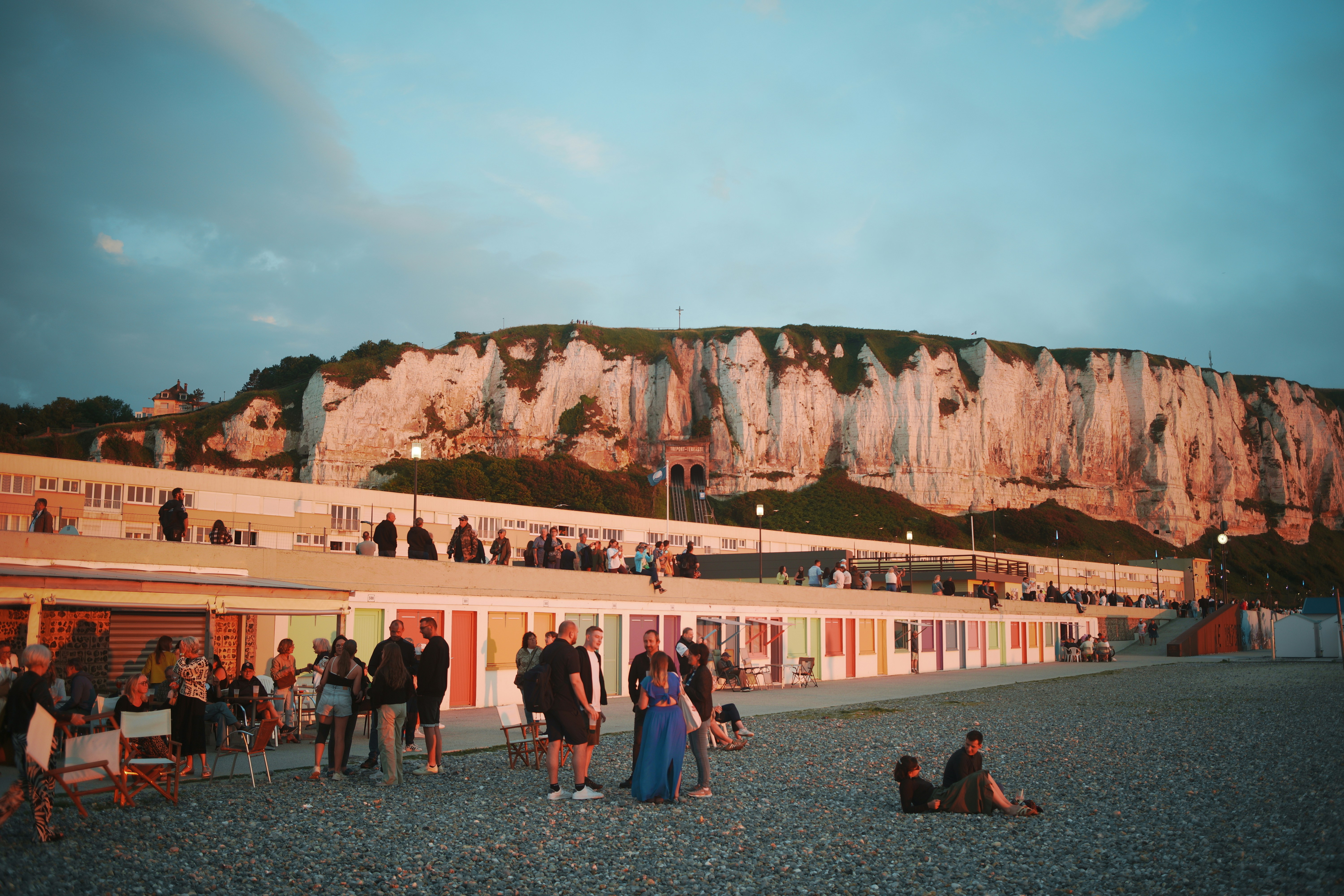 Crowd gathers along a seaside boardwalk with pastel market stalls and a rugged chalk cliff backdrop at dusk. A calm beach scene captured with a lively social atmosphere.