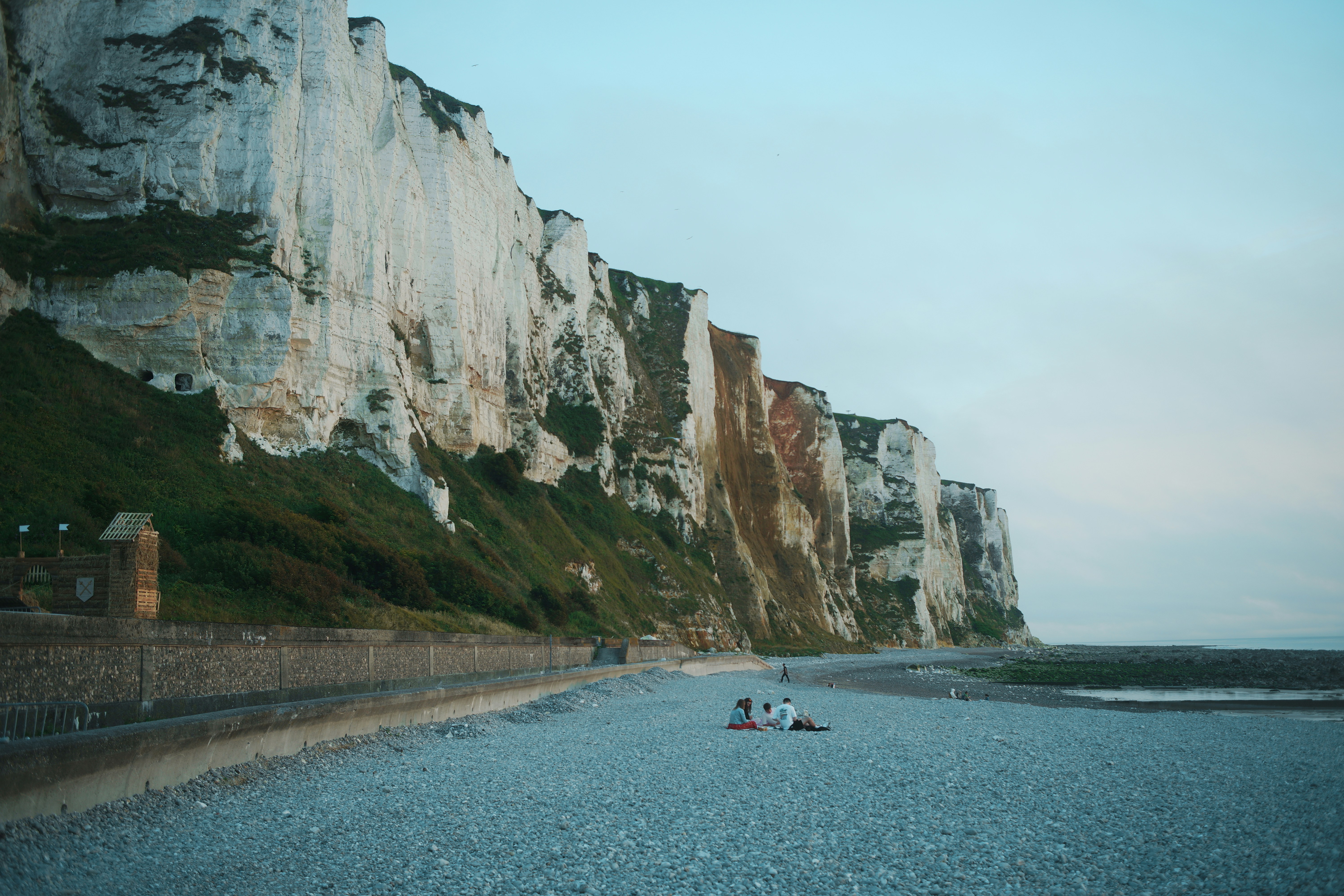 People sit on a pebble beach near towering white cliffs as the sea coolly meets the shore in a calm, overcast scene.