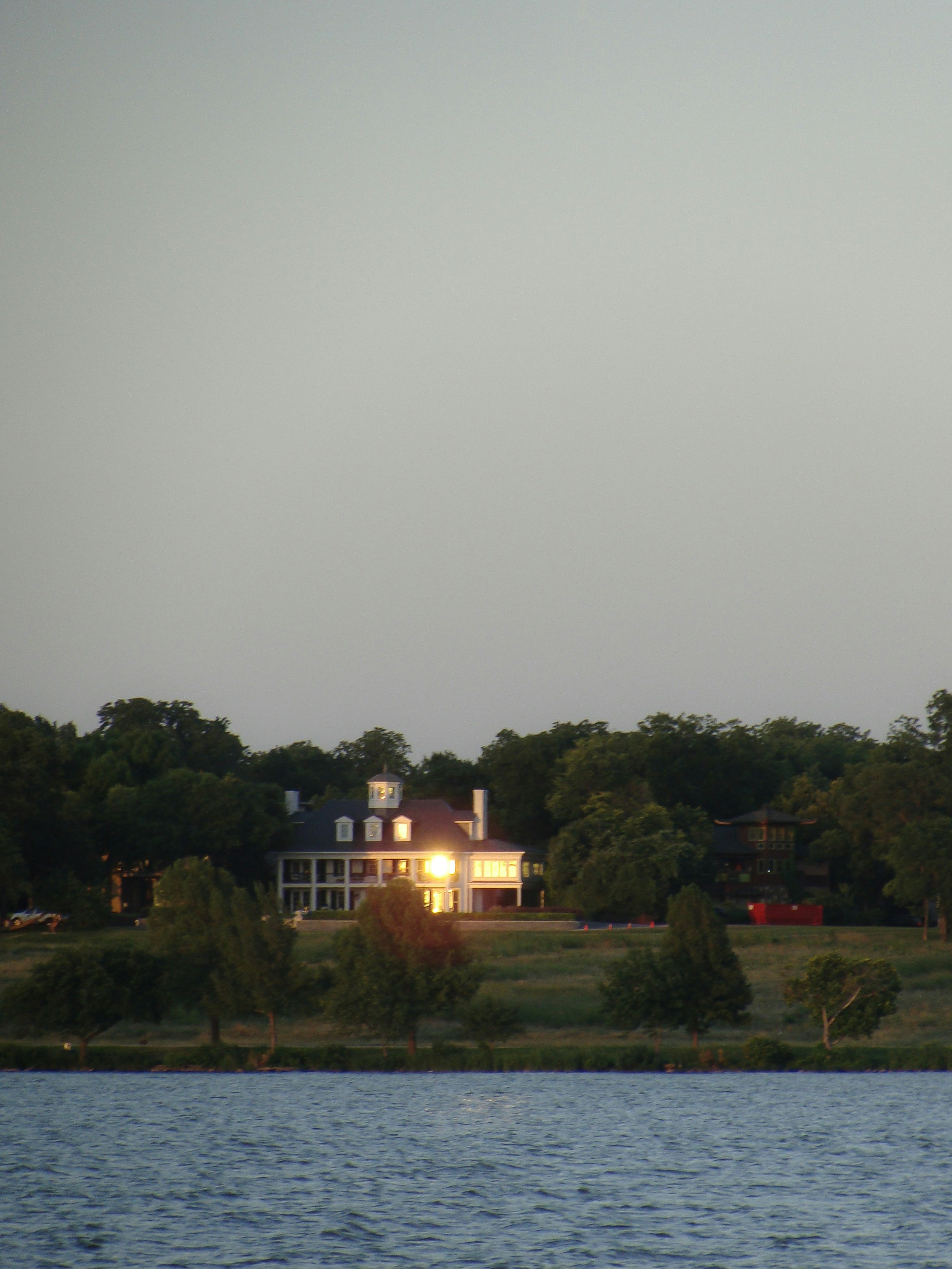 A lakeside house rests along a tree-lined shore as the last light glows on its facade, with calm water in the foreground and a pale sky above. Photograph captures a tranquil lakeside scene with warm building illumination.
