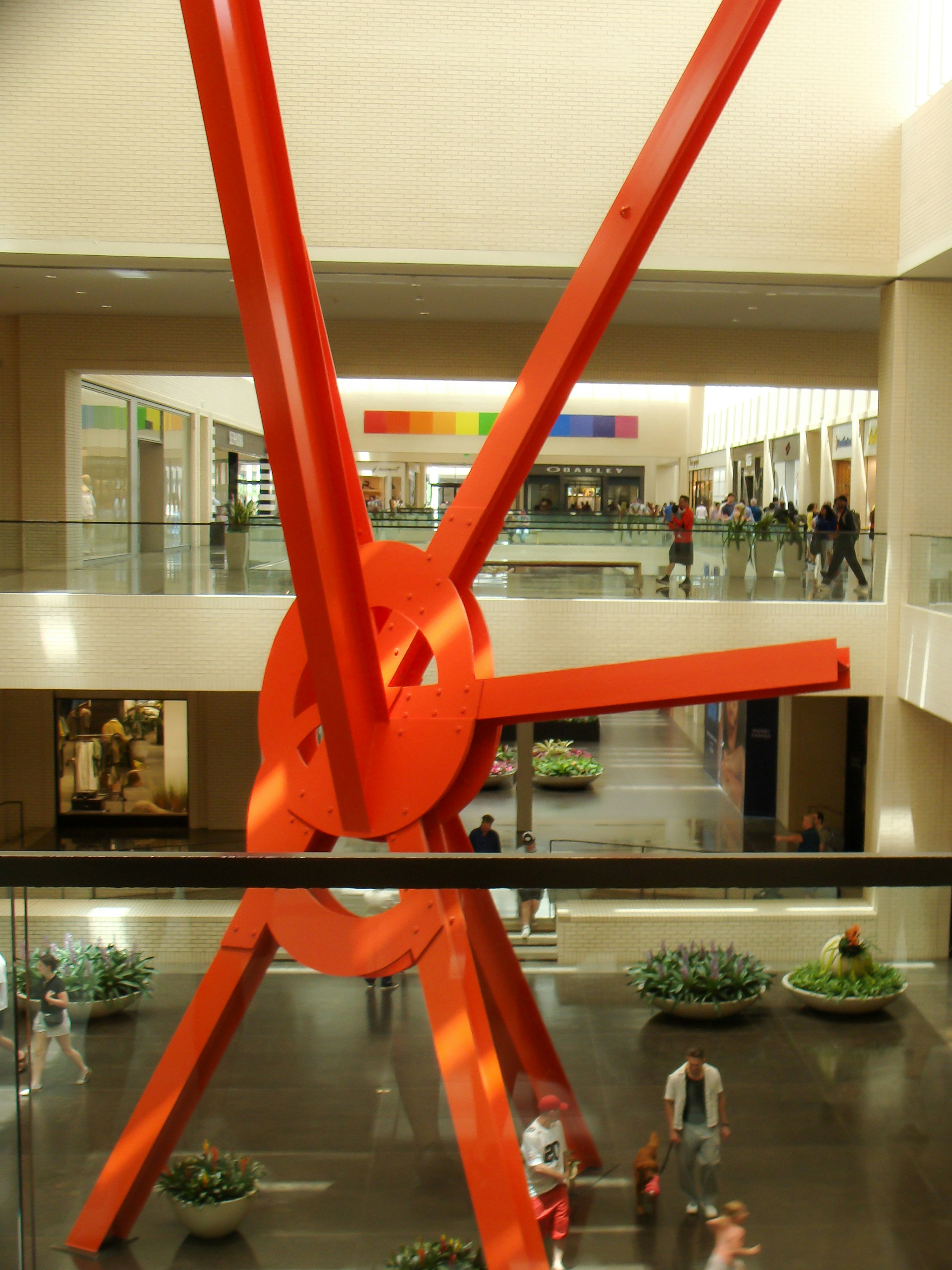 Bright red geometric sculpture dominates a busy mall atrium, with shoppers and storefronts visible in the background; the composition emphasizes architectural contrast and crowds around the art piece.
