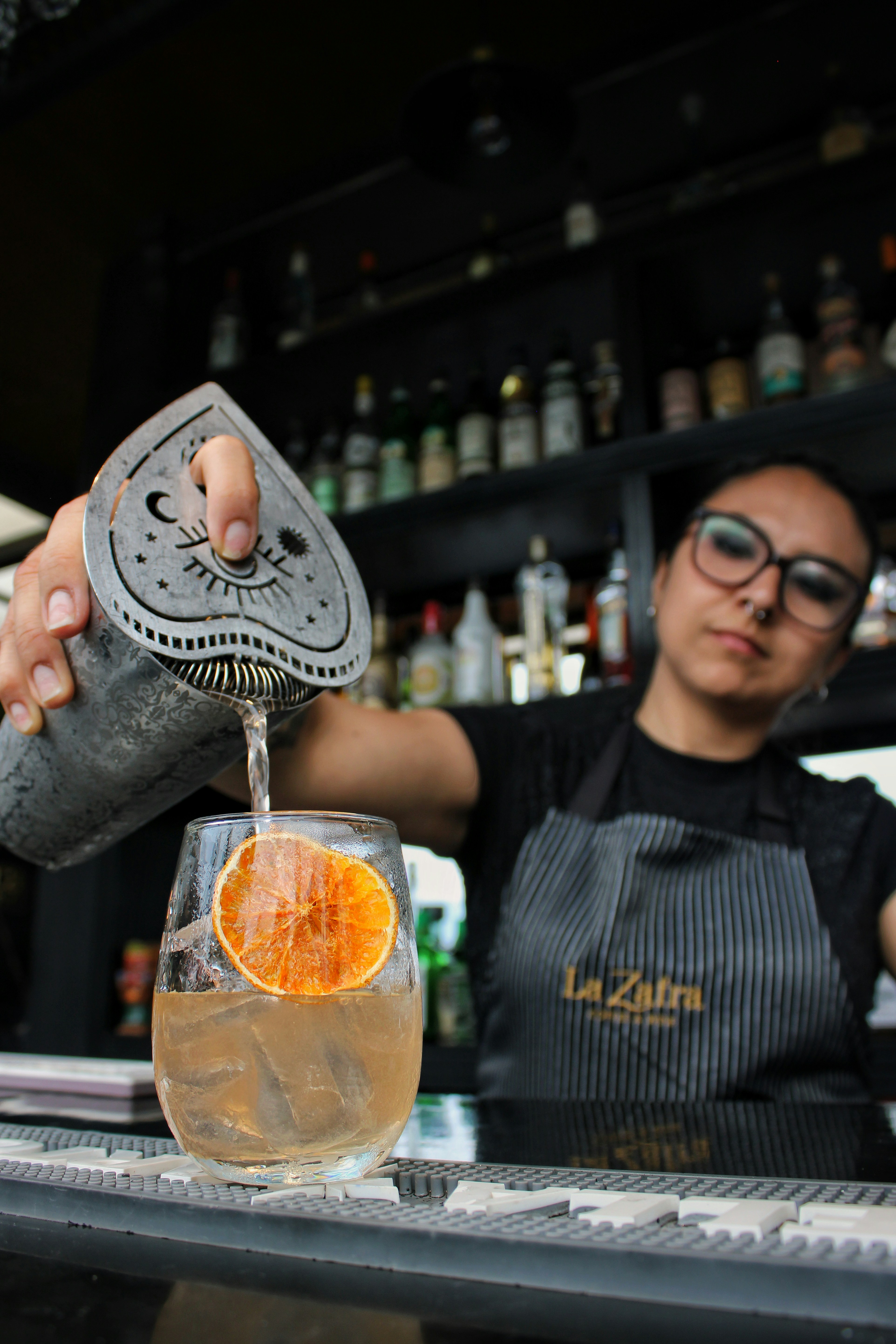 Bartender pours cocktail with orange slice from decorative shaker.