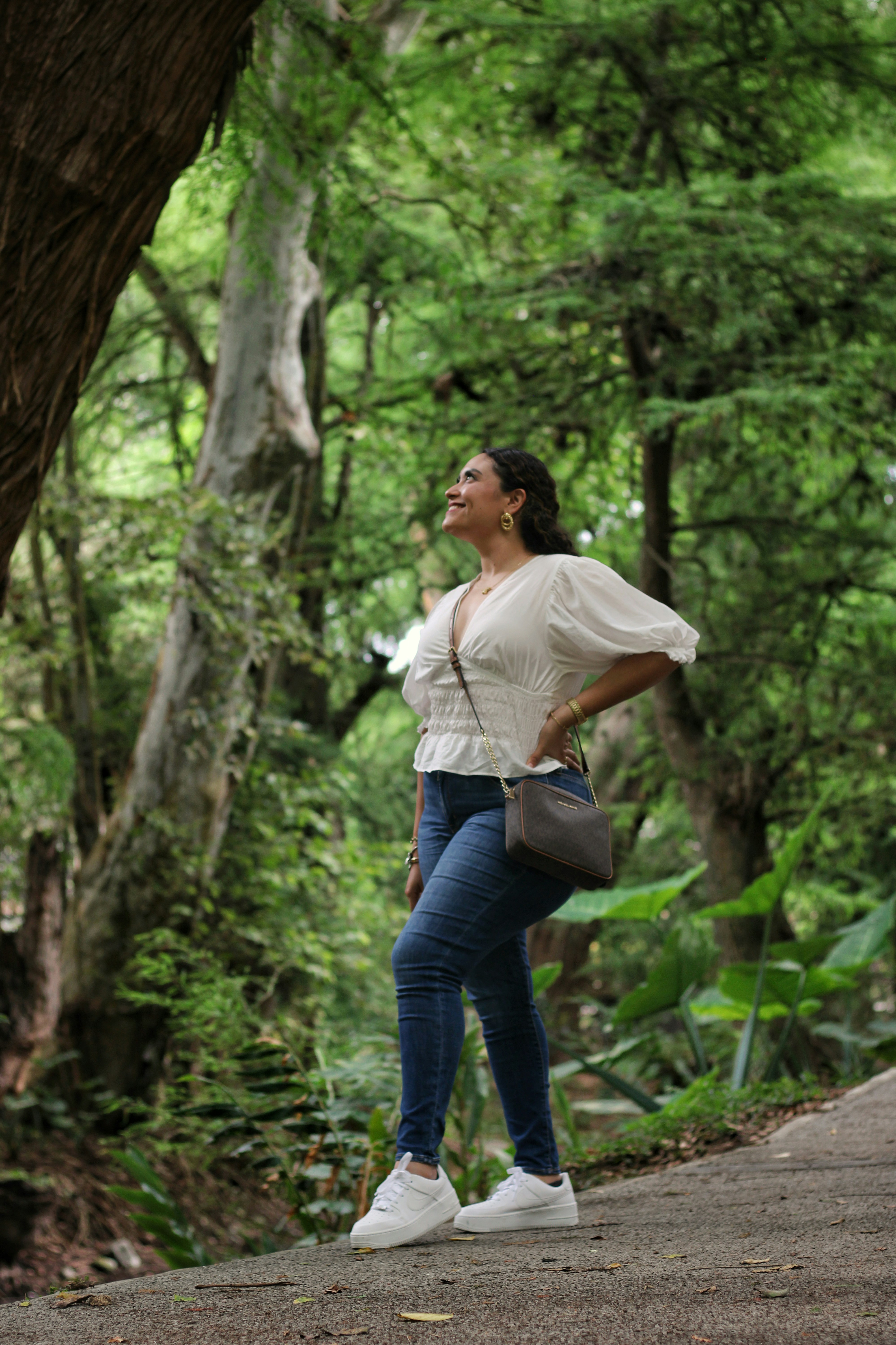 A woman stands on a forest path, looking upward with a smile, wearing a white blouse, jeans, and white sneakers; a crossbody bag hangs at her side amid lush trees. The scene emphasizes her relaxed pose and natural surroundings.
