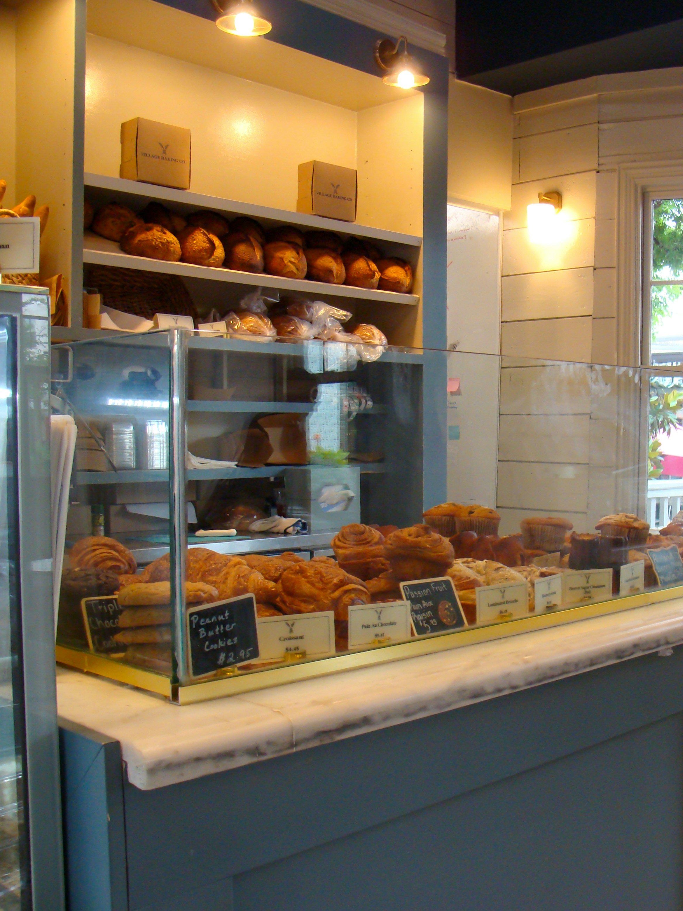 Assorted pastries showcased behind glass, with shelves of bread and boxes in the background, inside a cozy bakery.