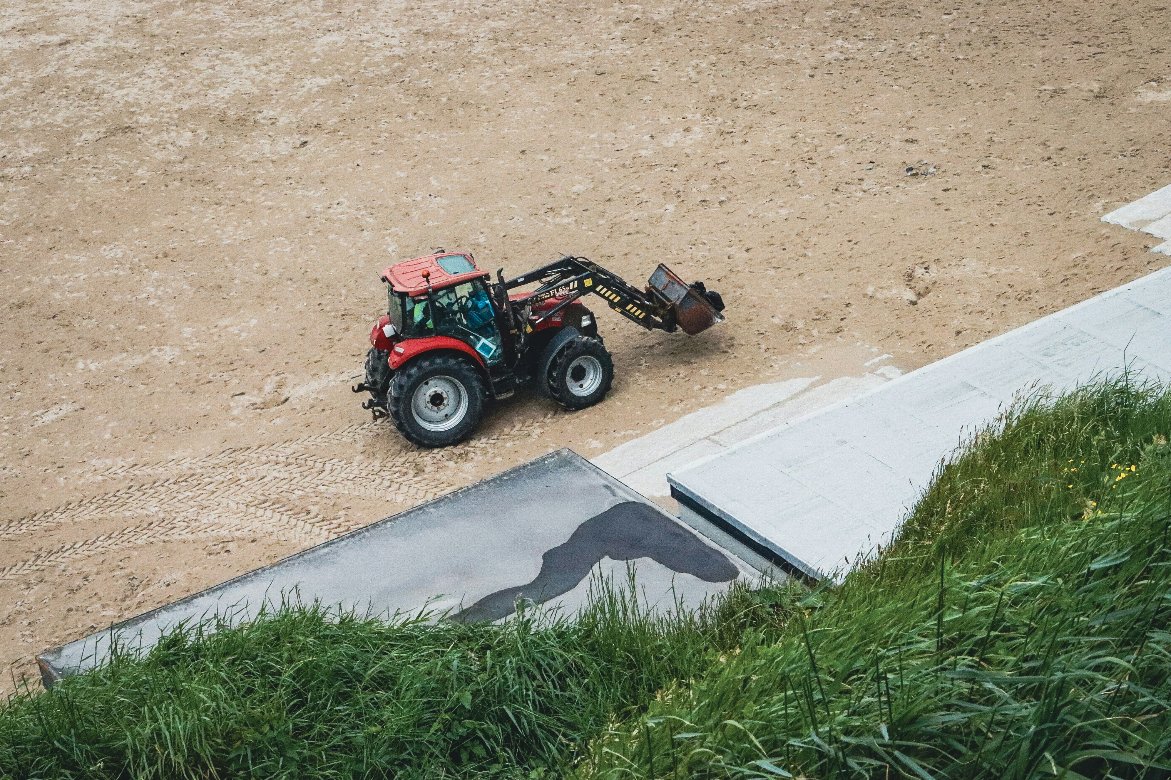 Red utility tractor with front loader parked on excavated dirt near a concrete ramp, viewed from above with green grass edge in the foreground. A construction site scene showing machinery and exposed soil.